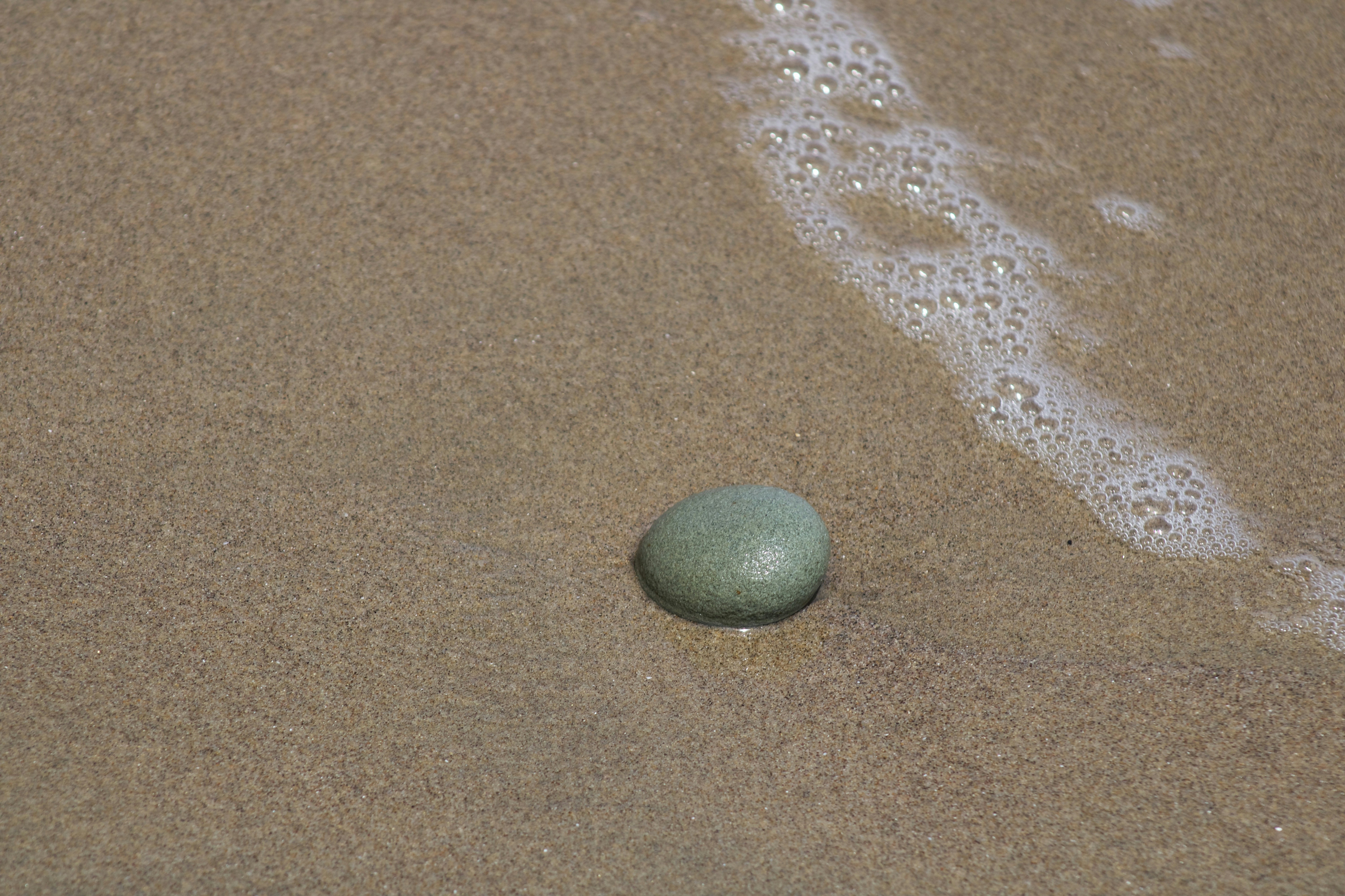 Smooth green stone resting on wet sand near the ocean's edge, with gentle waves lapping nearby.