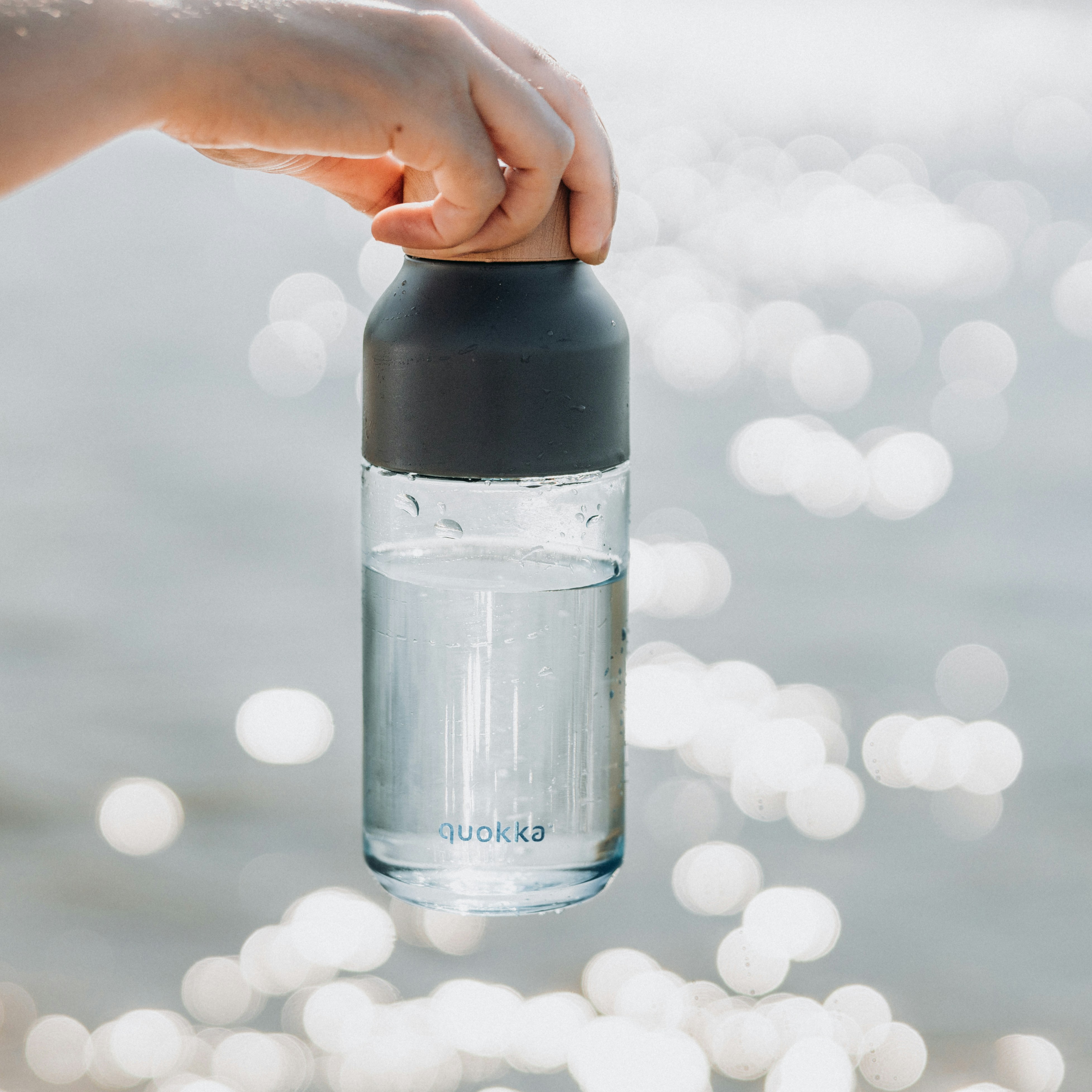 A hand holding a clear water bottle against a shimmering backdrop of sunlight reflecting on water. The bottle features a sleek design and a logo.