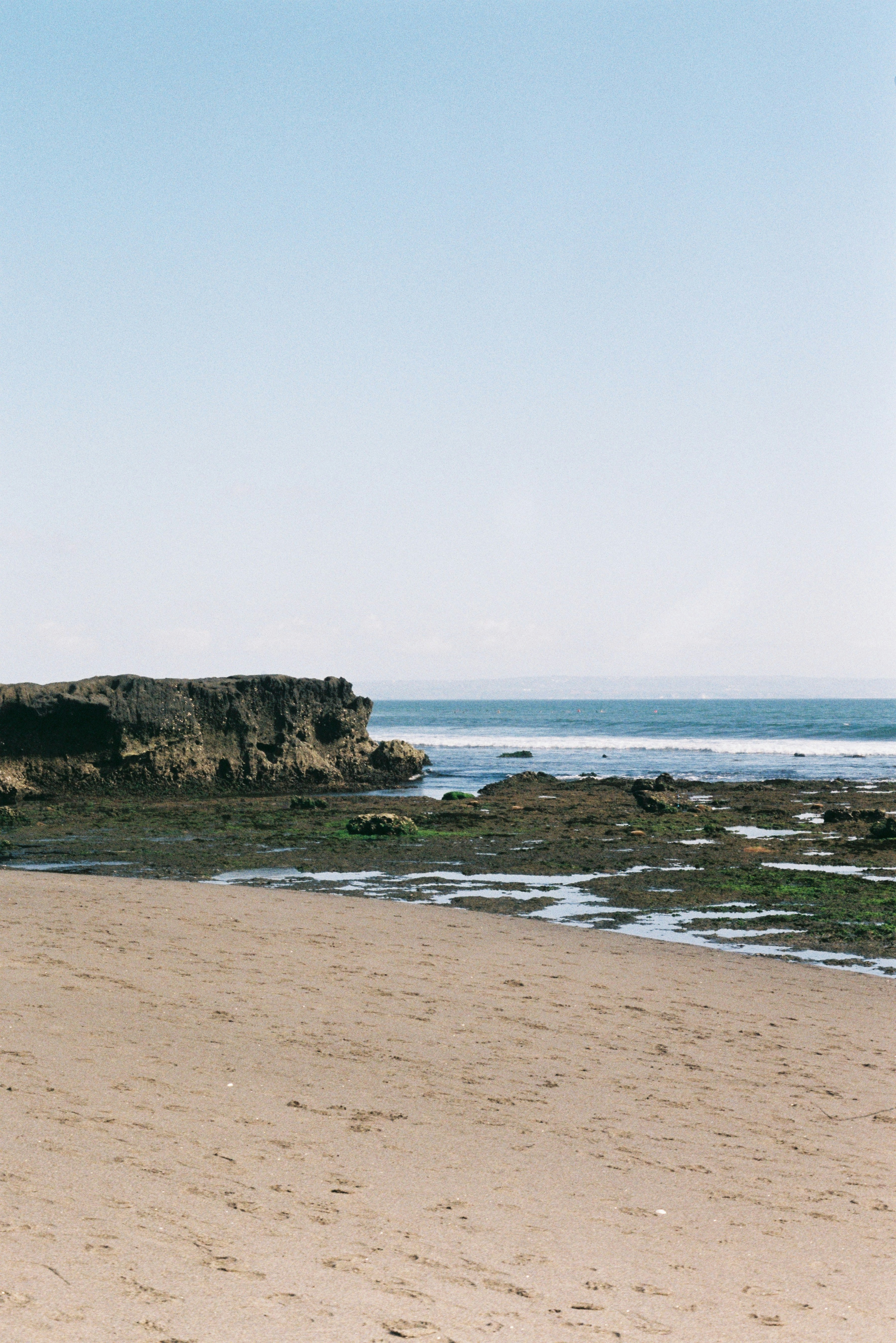 A tranquil beach scene with a rocky outcrop along the shore, shallow tidal pools, and a calm horizon over the sea.
