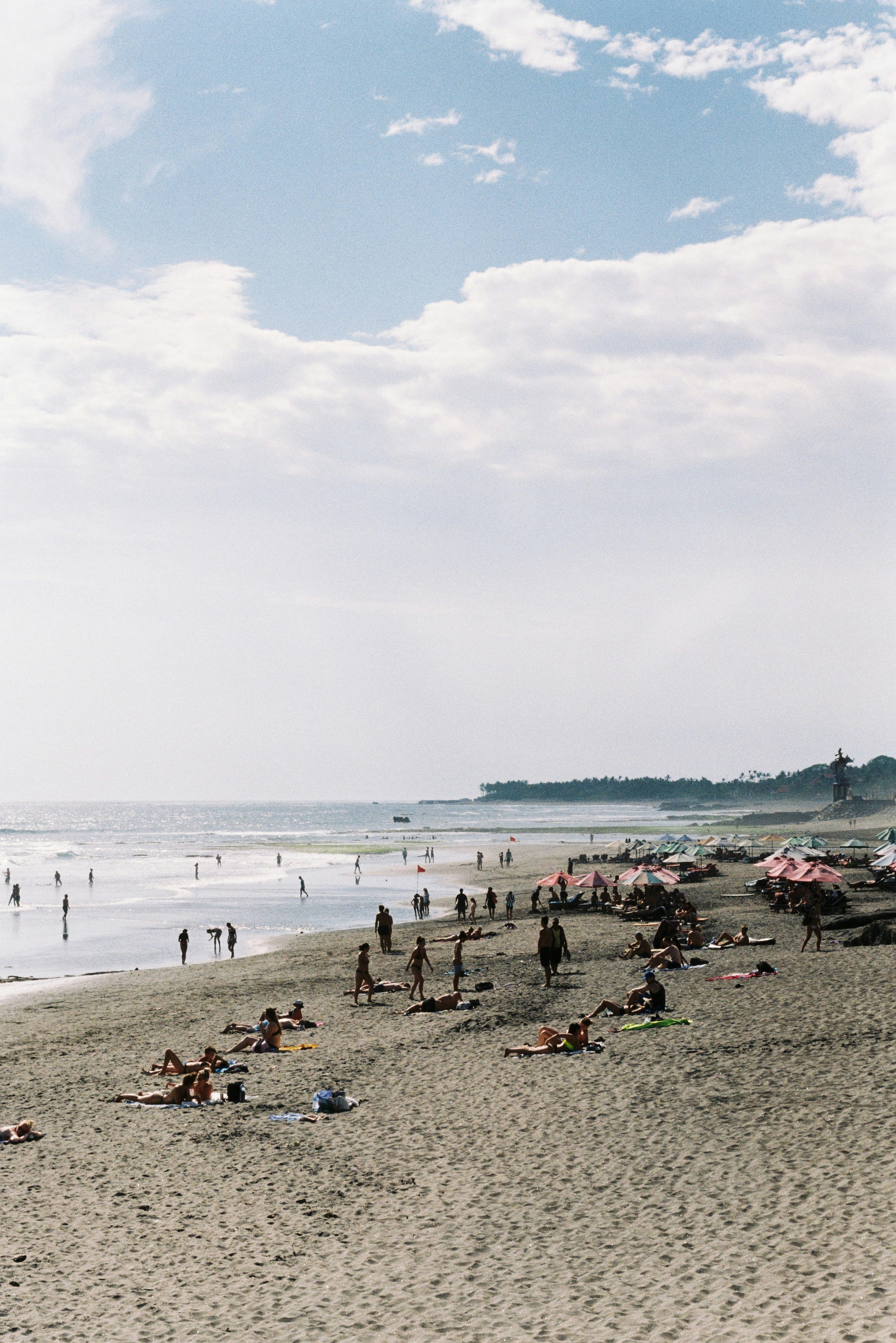 Vibrant beach scene with sunbathers and surfers enjoying the waves under a partly cloudy sky.