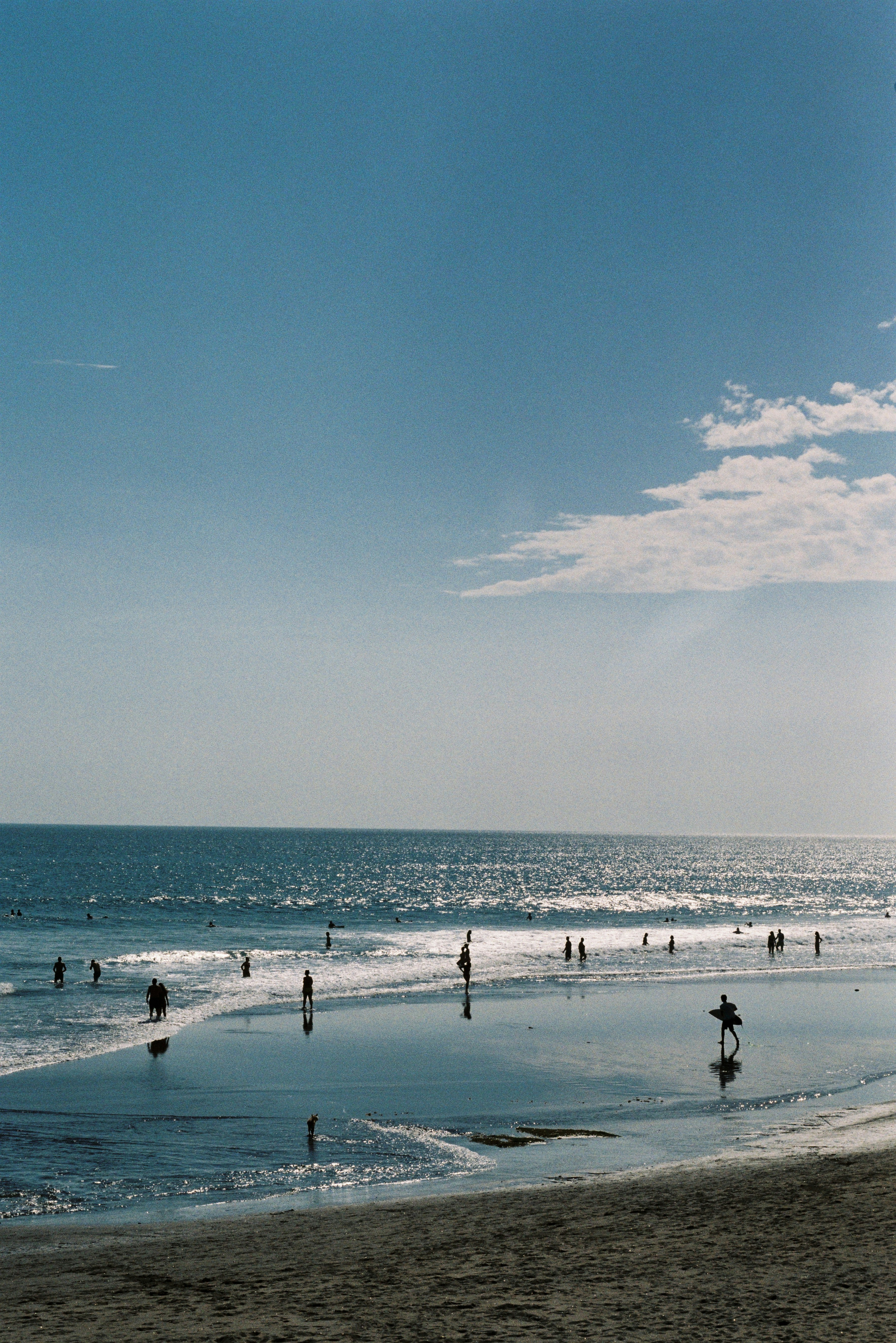 Beachgoers enjoying the gentle waves under a clear blue sky, with surfers riding the surf in the distance.