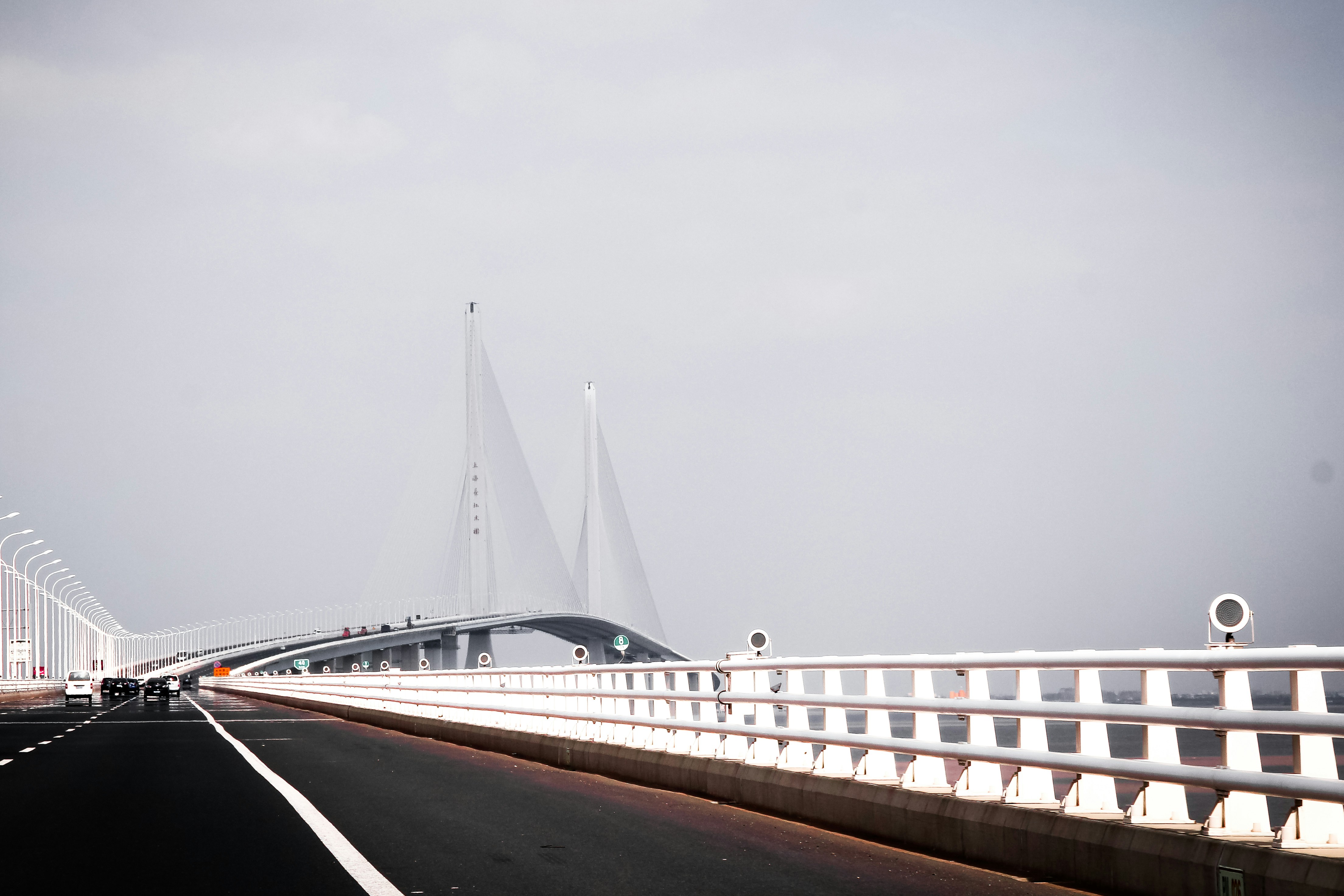 A modern suspension bridge stretches across a highway, enveloped in a soft haze, showcasing its elegant architecture against a muted sky.