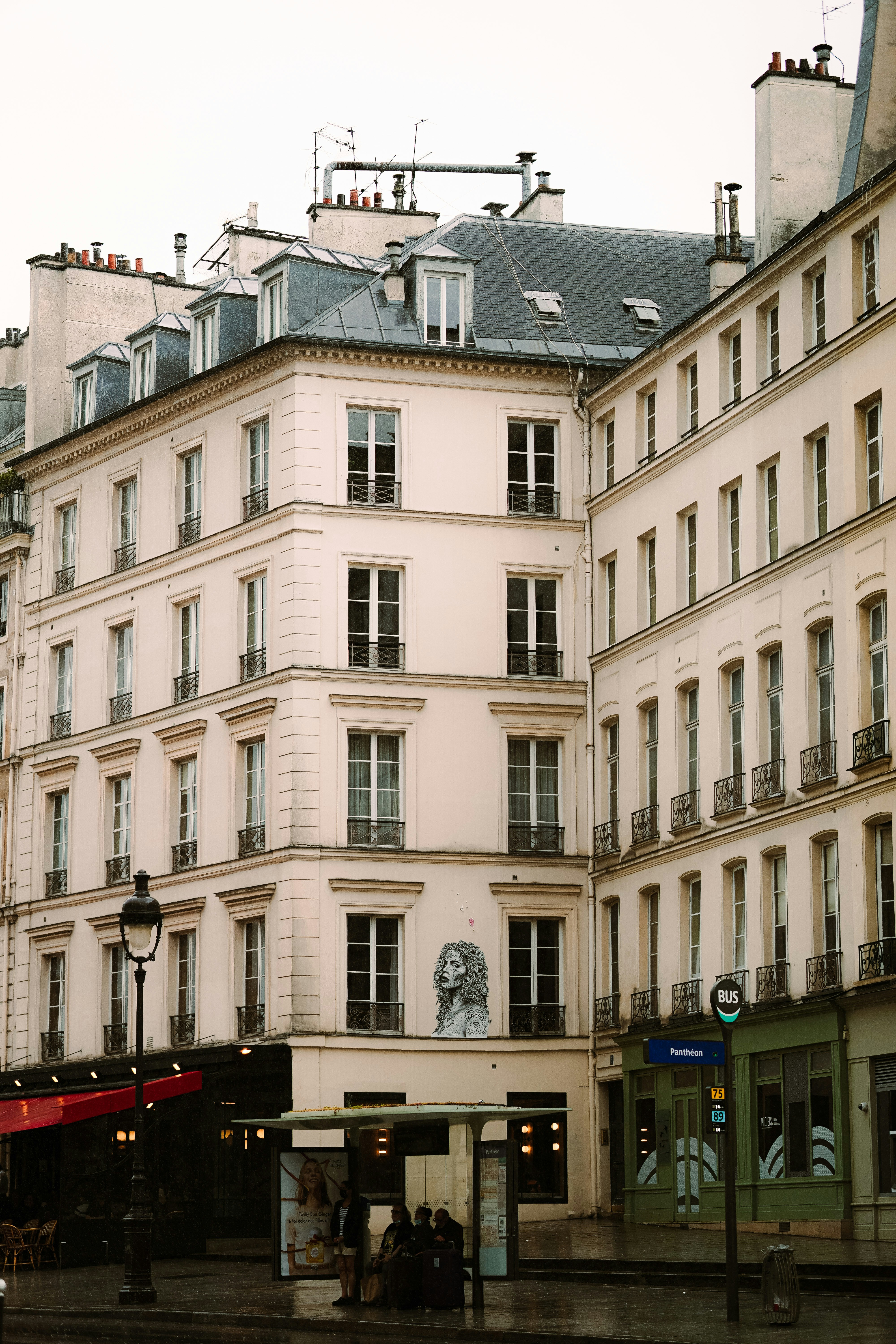 Elegant Parisian buildings with intricate details and a street-side bus stop create a serene urban scene. A mural adds a touch of artistic flair.