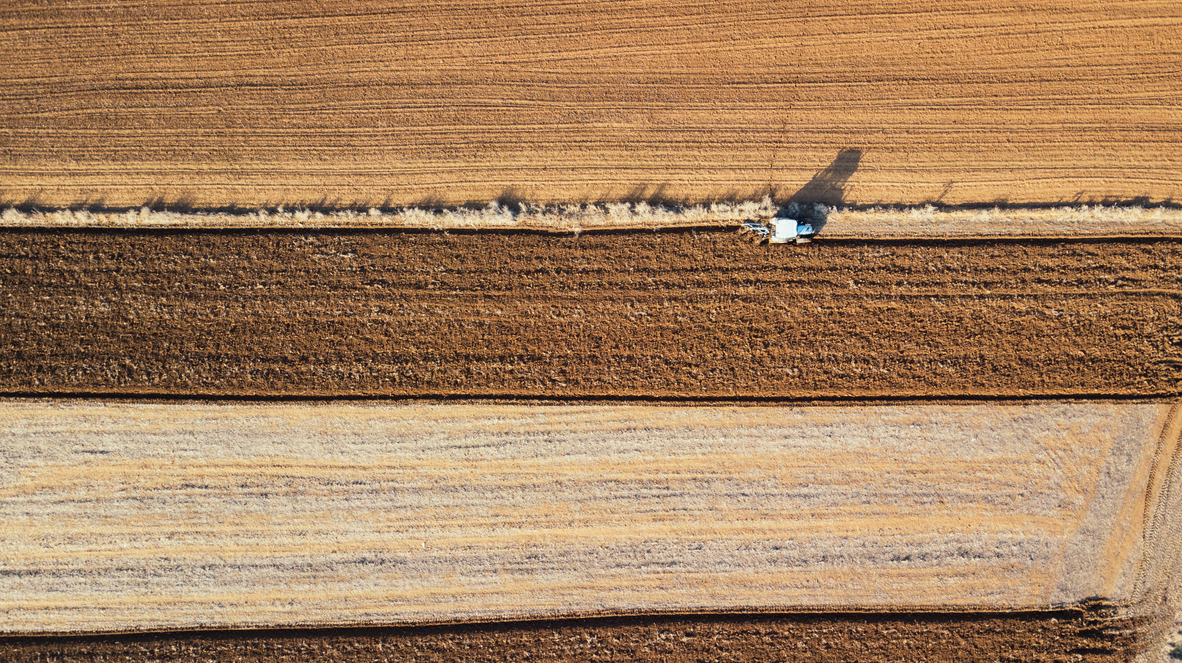 Aerial view of a tractor working across meticulously arranged fields, showcasing the intricate patterns of cultivated land. The contrast of earthy tones highlights the agricultural landscape.