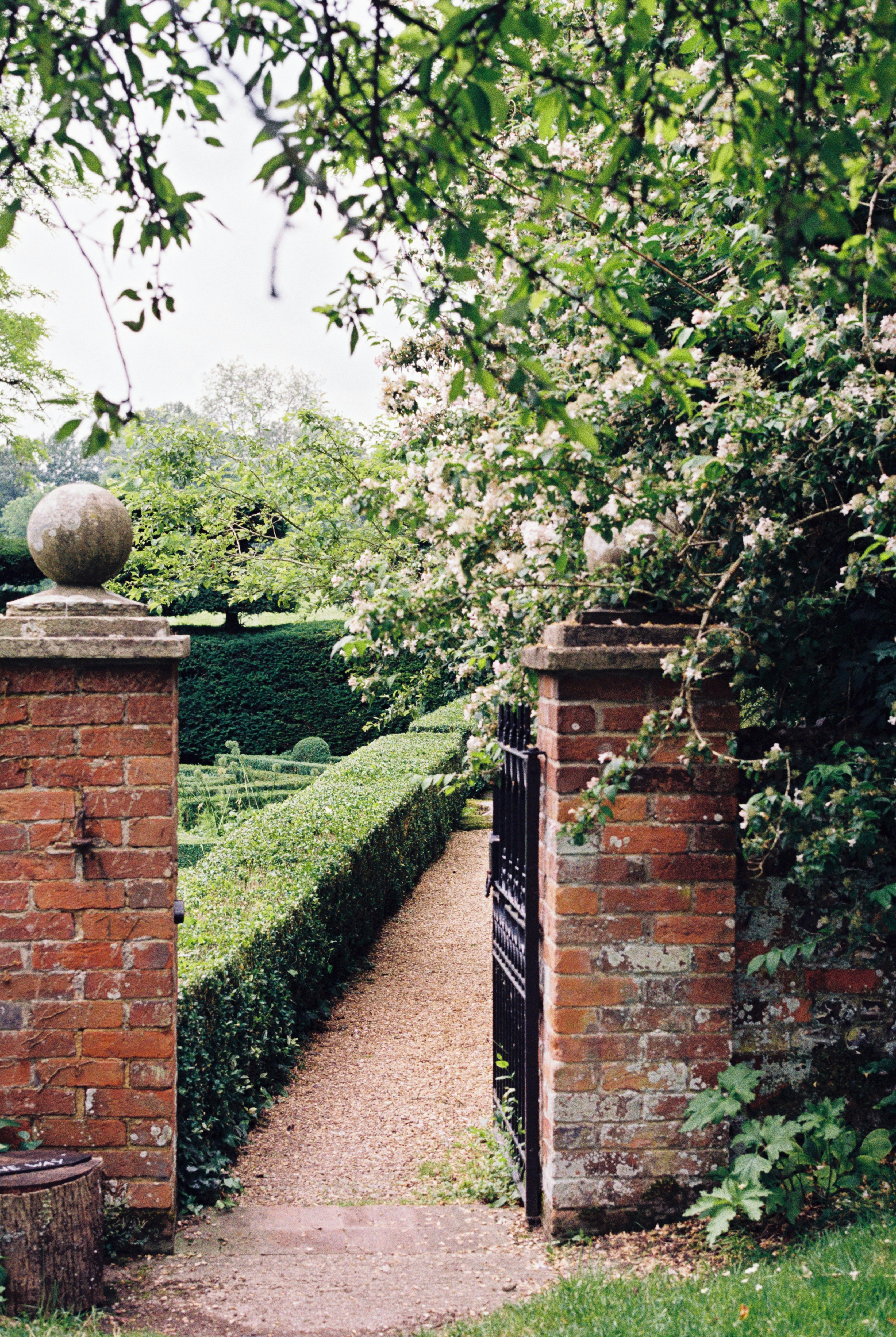 A serene pathway framed by brick pillars and lush greenery, leading into a meticulously manicured garden. The entrance is adorned with a spherical finial atop the gateposts.