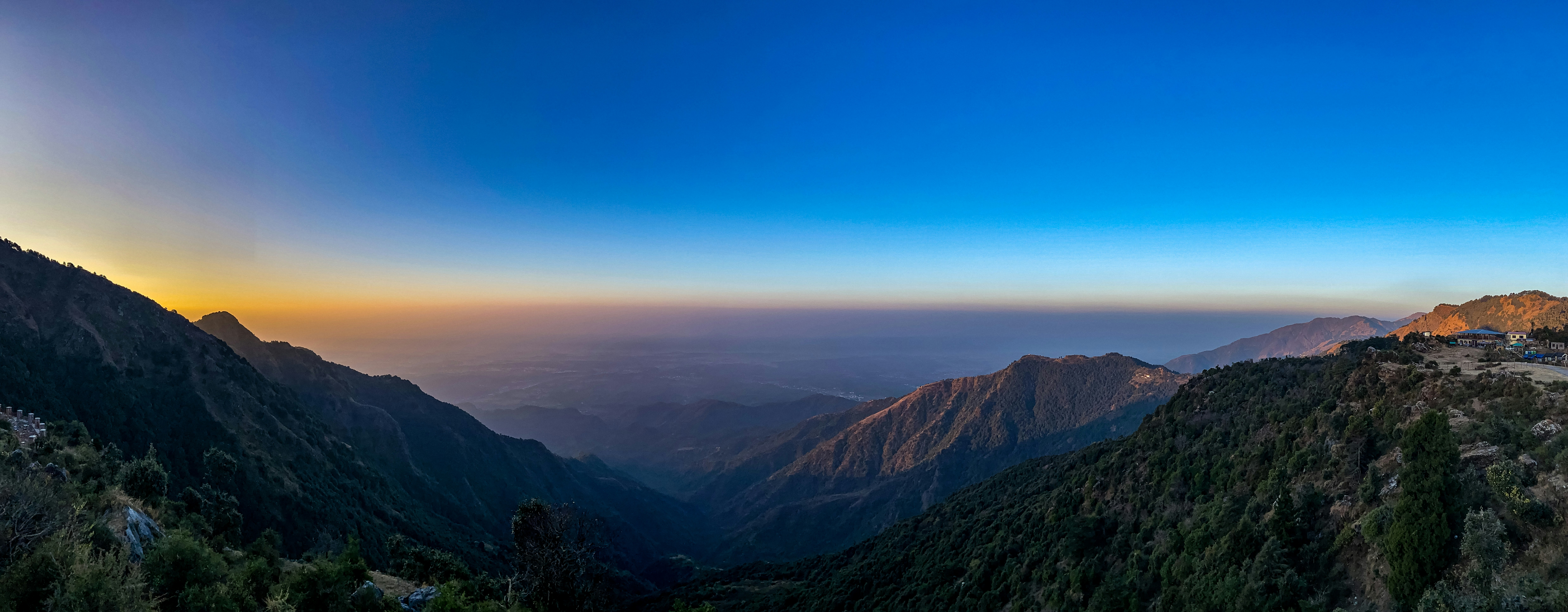 Panoramic view of a mountain landscape at dusk, showcasing a gradient of colors from warm to cool tones across the sky. The vastness of the valley below adds depth to the scene.