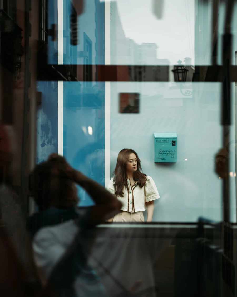 A woman stands against a pale blue wall beside a turquoise mailbox, seen through layered reflections and dark window frames. The composition feels urban and contemplative.