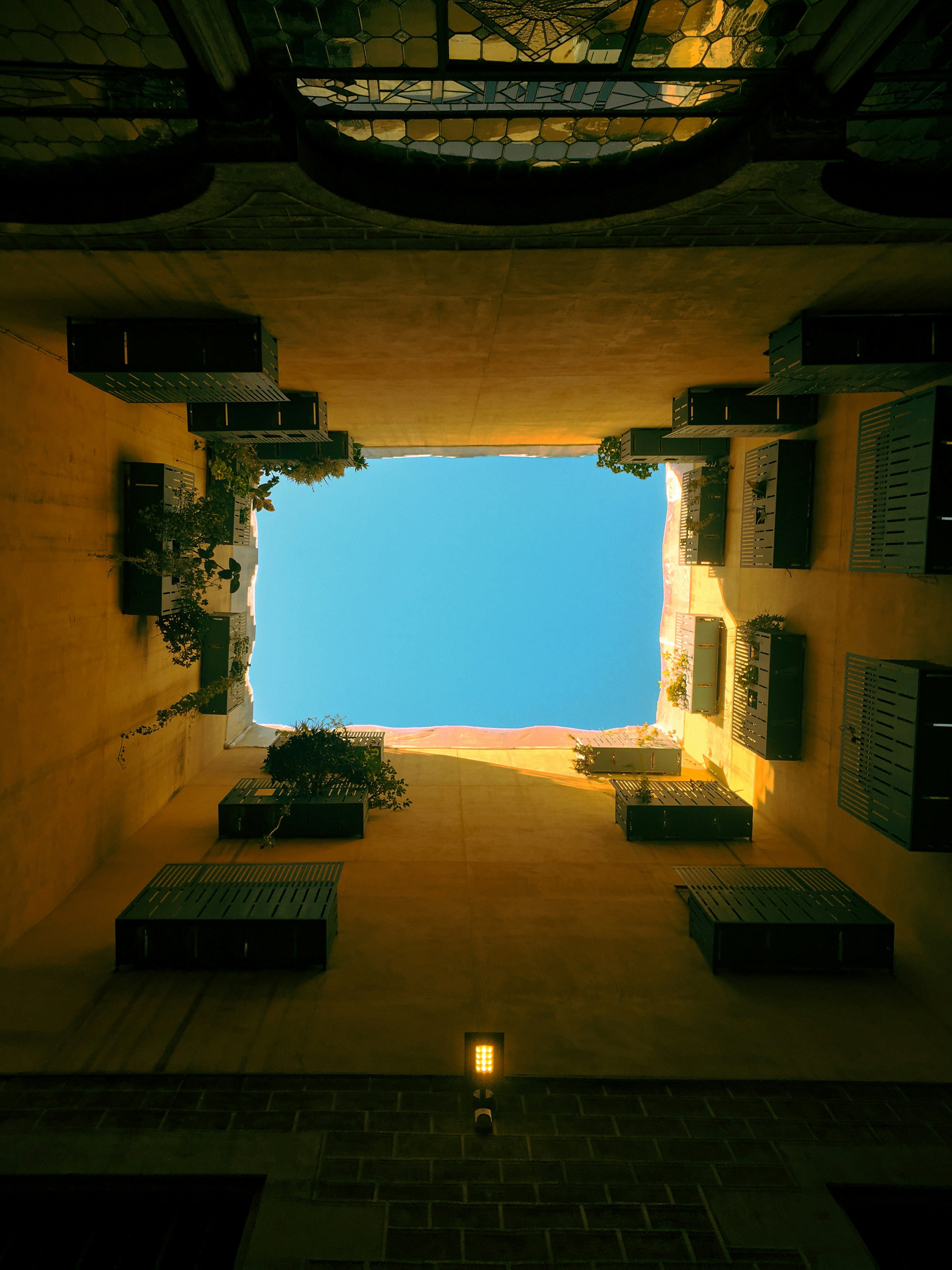 View from below of a courtyard surrounded by buildings, framed by vibrant blue sky and greenery. The architectural details and light create a unique atmosphere.