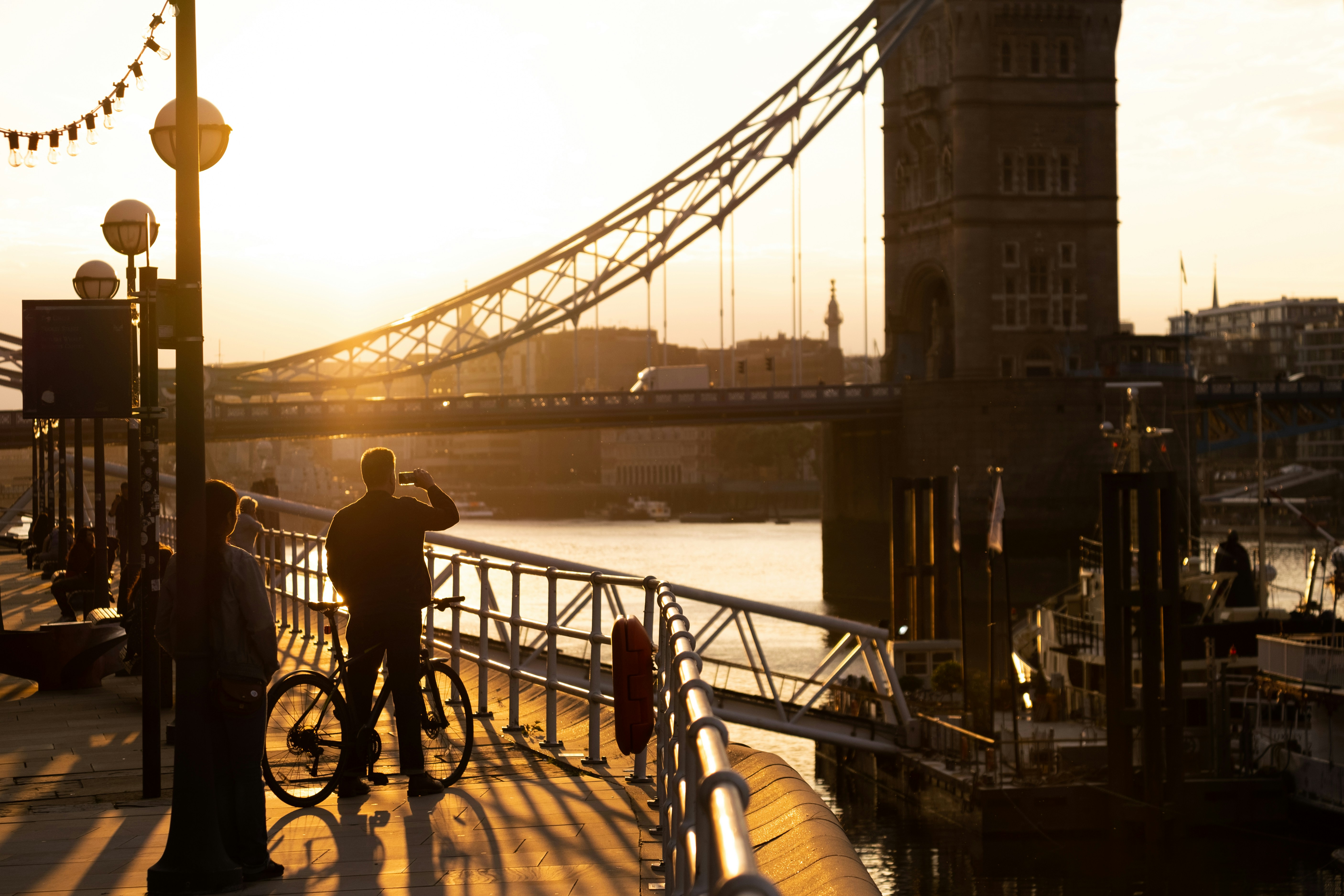 This captivating image captures a serene moment by a bridge at sunset, where a silhouetted figure stands with a bicycle, capturing the scene on a phone. The warm golden light bathes the area, casting long shadows and highlighting the intricate details of the bridge structure against a soft, glowing sky. The juxtaposition of modern life and historical architecture creates a visually striking and atmospheric composition.