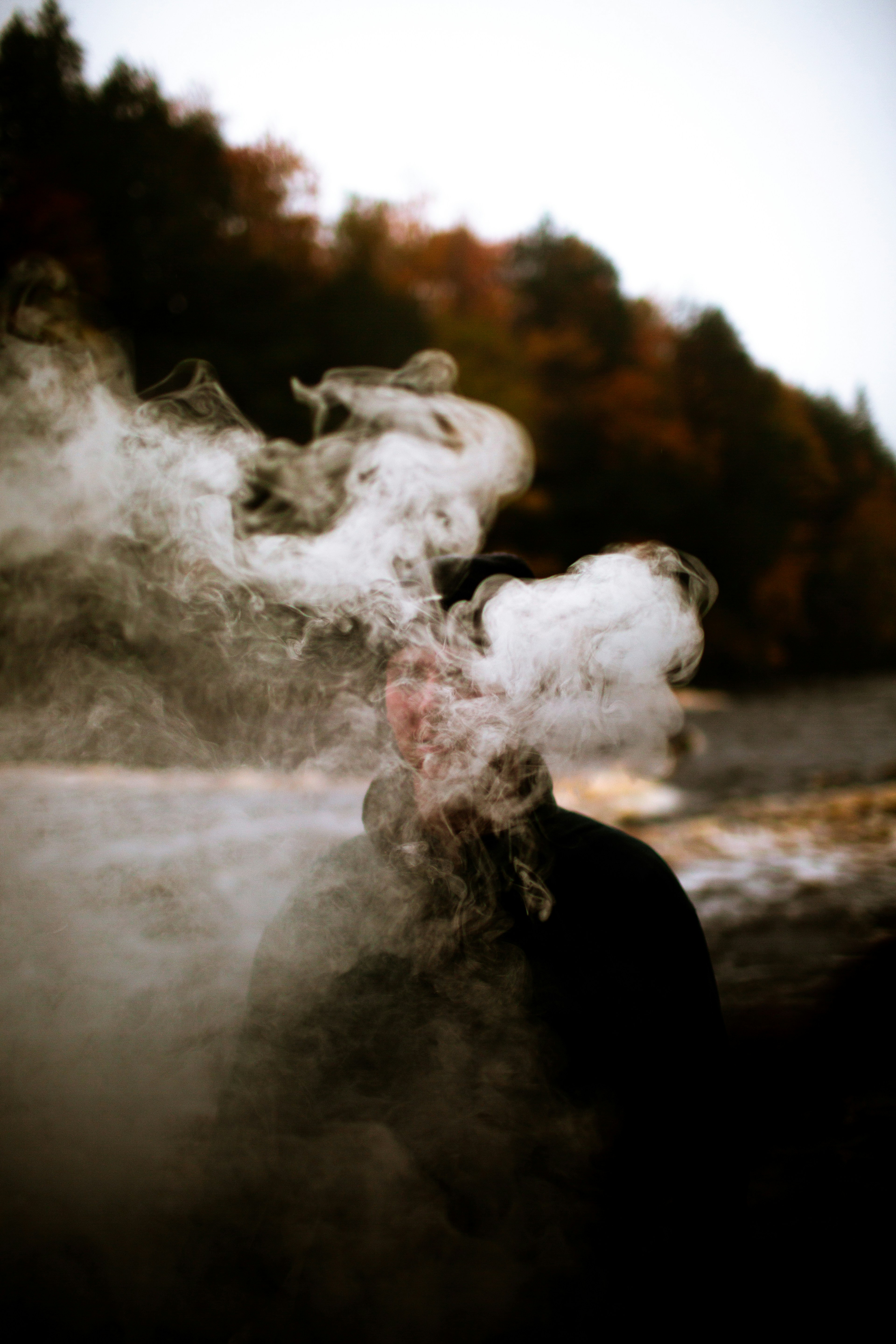 A figure shrouded in swirling vapor stands by a river, surrounded by autumn foliage. The ethereal smoke creates a dreamlike atmosphere.
