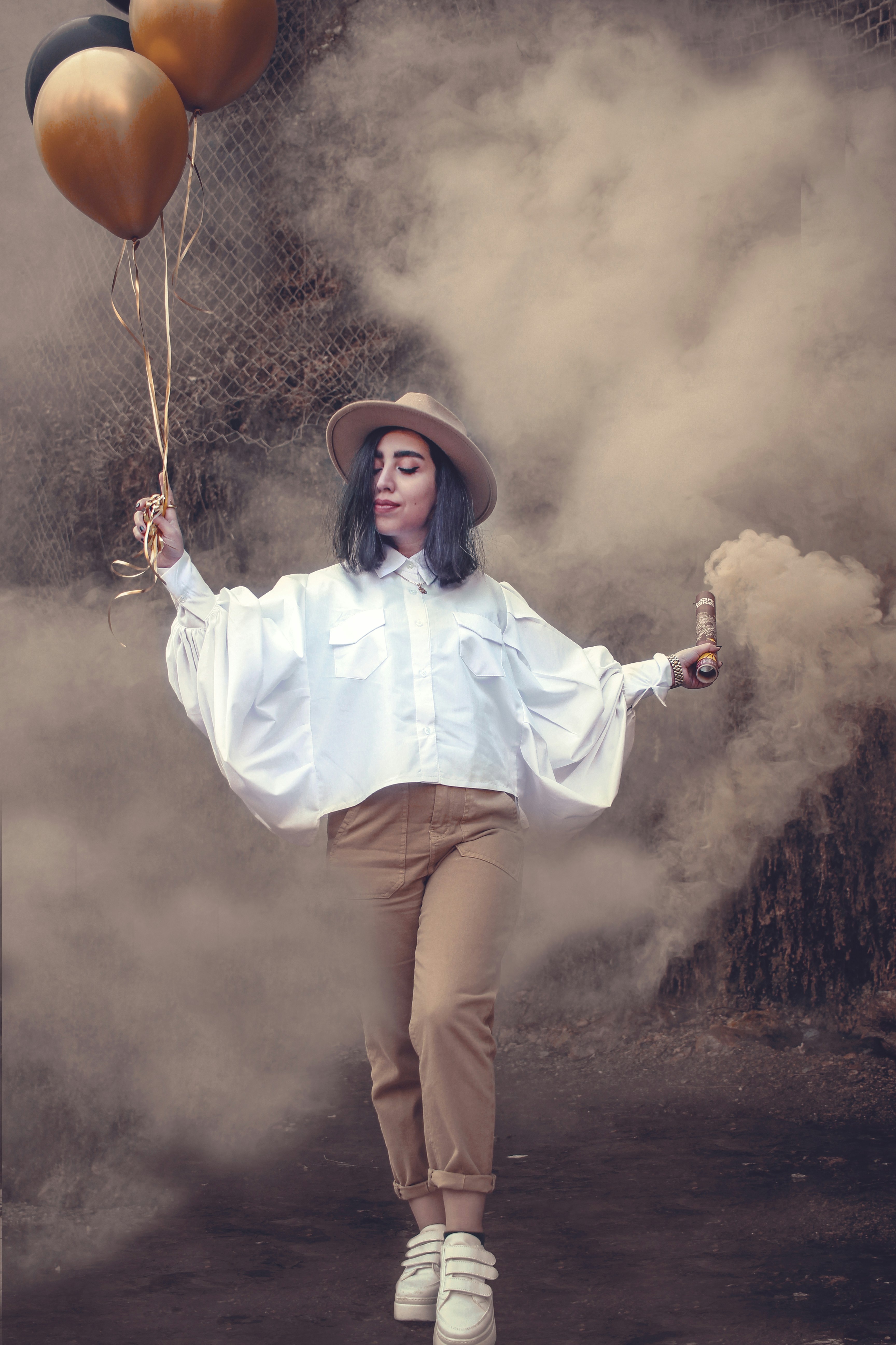 A woman in a flowing white shirt and beige pants holds black and gold balloons while releasing colorful smoke, creating a whimsical atmosphere.