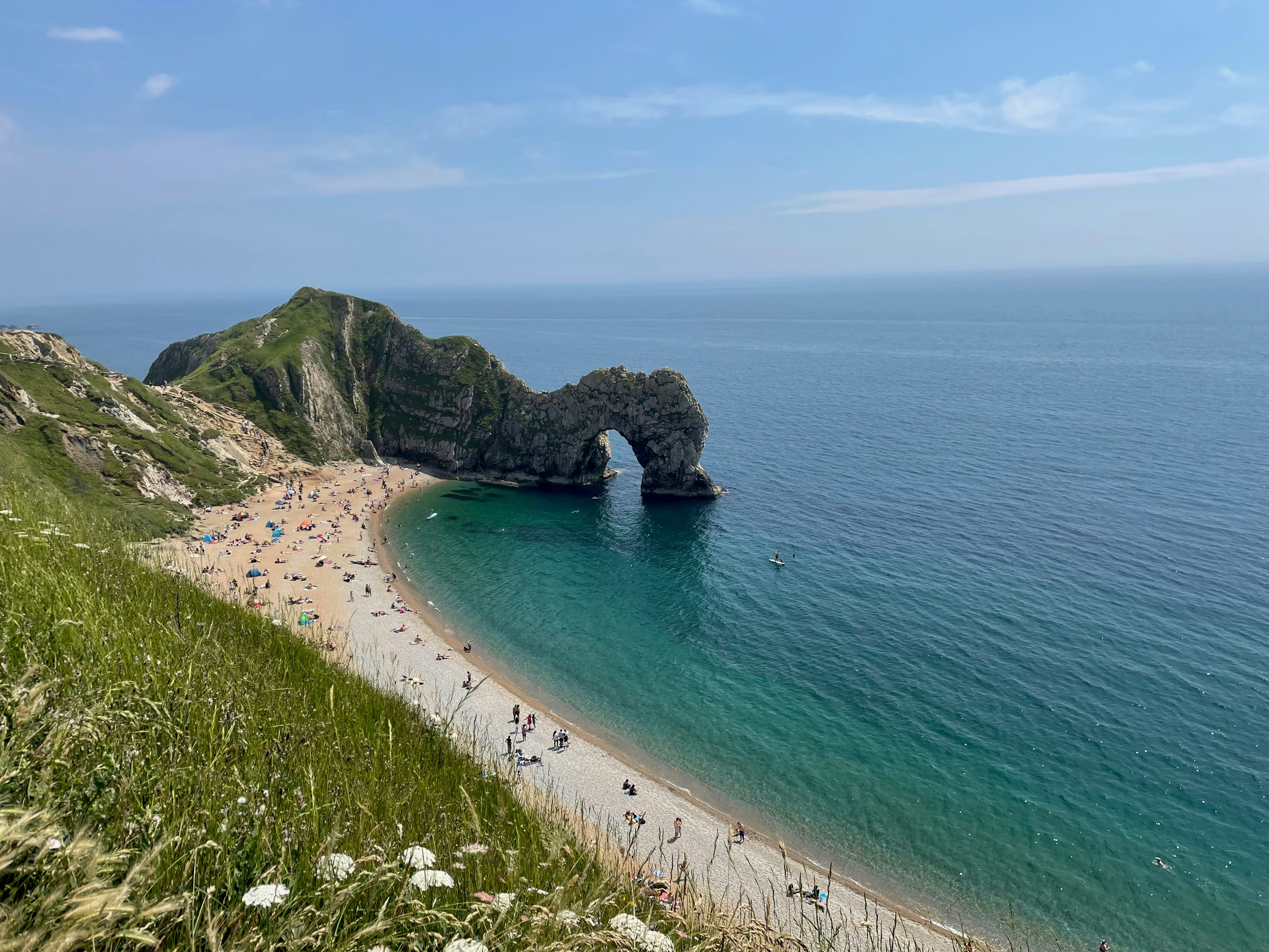 Durdle Door rock formation stands majestically over a sandy beach, with sunbathers enjoying the serene coastal landscape. The turquoise waters invite exploration.