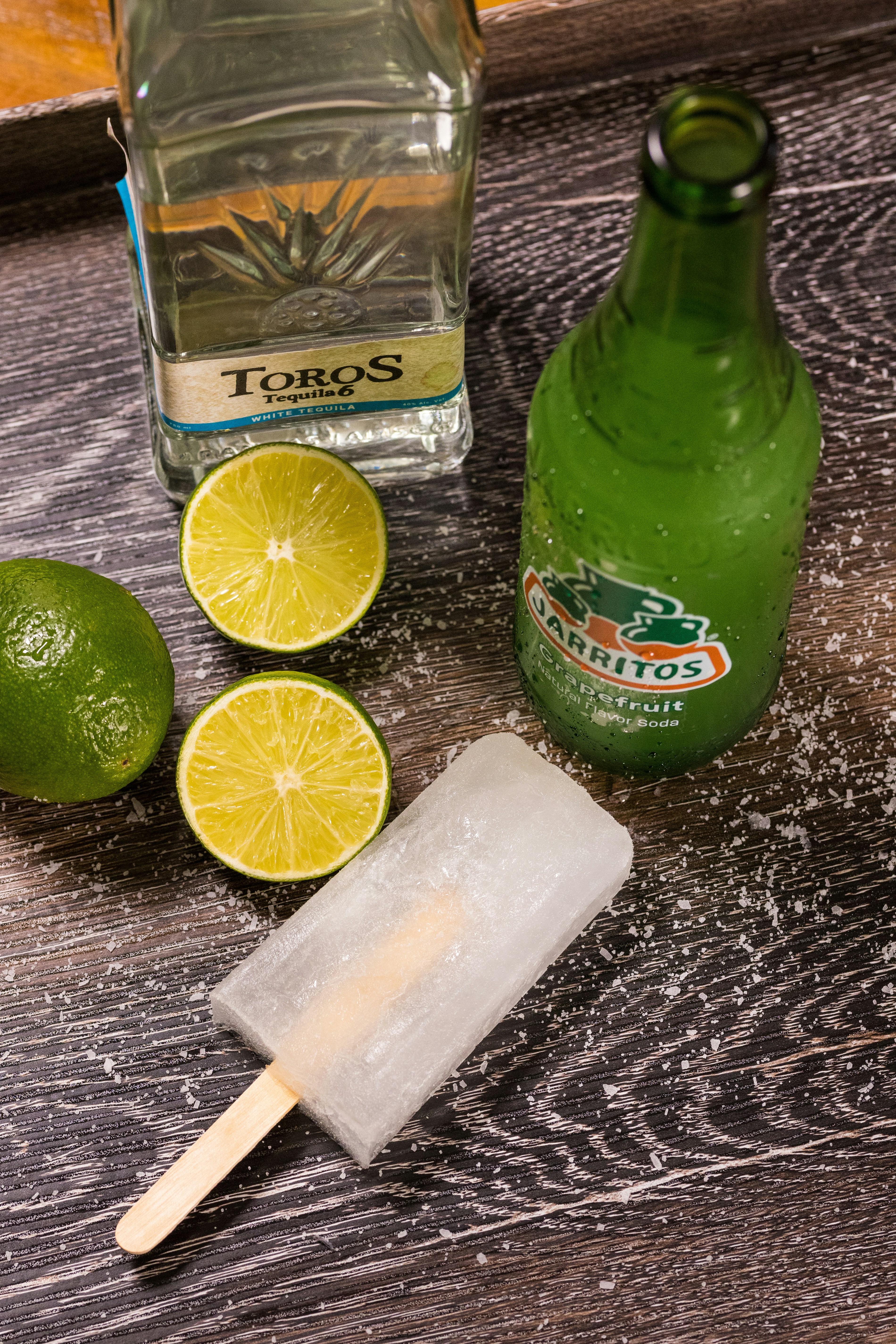 Tequila bottle, lime halves, and a frosty popsicle beside a green soda on a wooden surface.