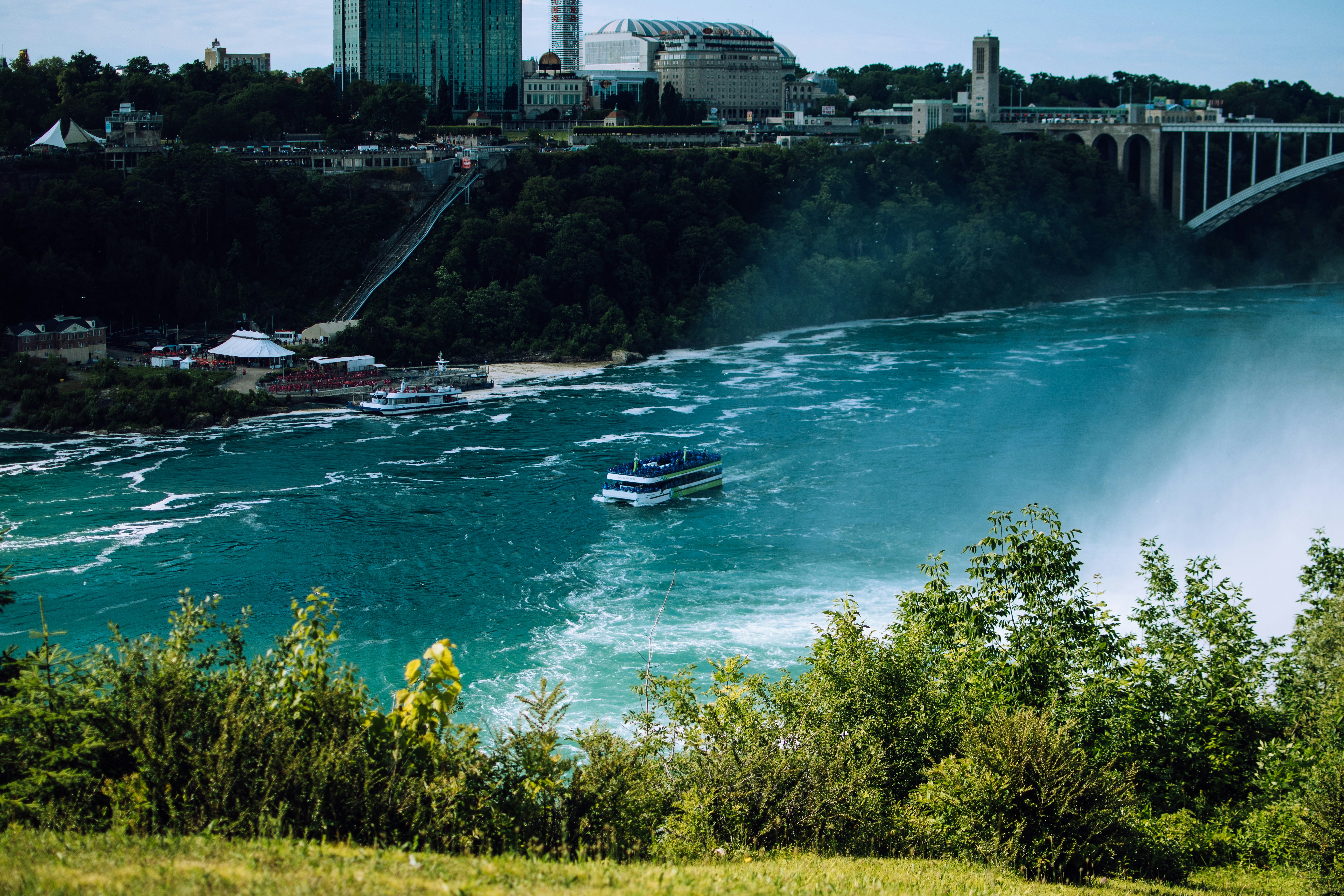 Tour boat navigating the turquoise waters near a cascading waterfall, with a lush green foreground and urban skyline in the background.