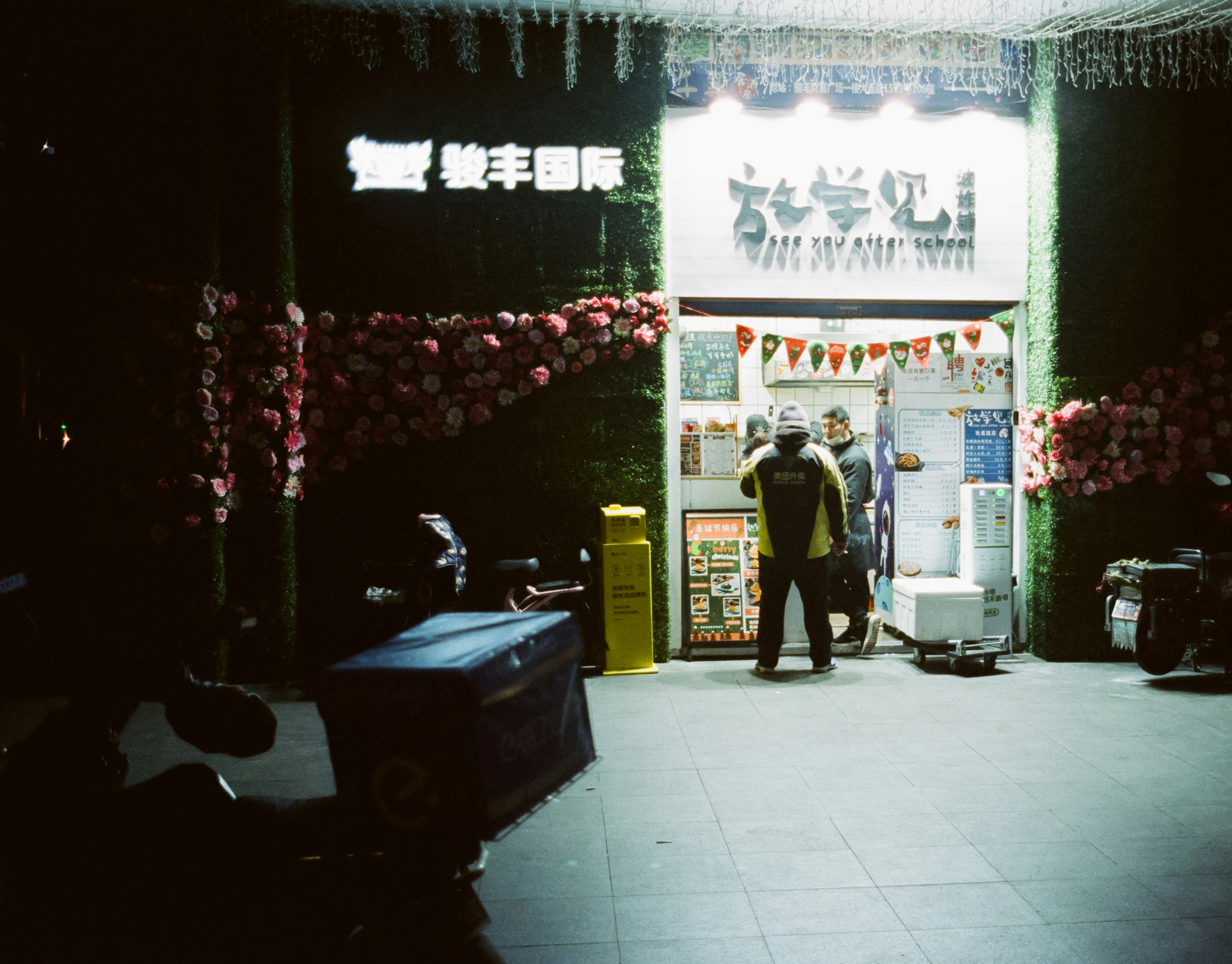Nighttime storefront entrance framed by floral walls and string lights. Two figures stand at the doorway under bright signage in a street photography scene.