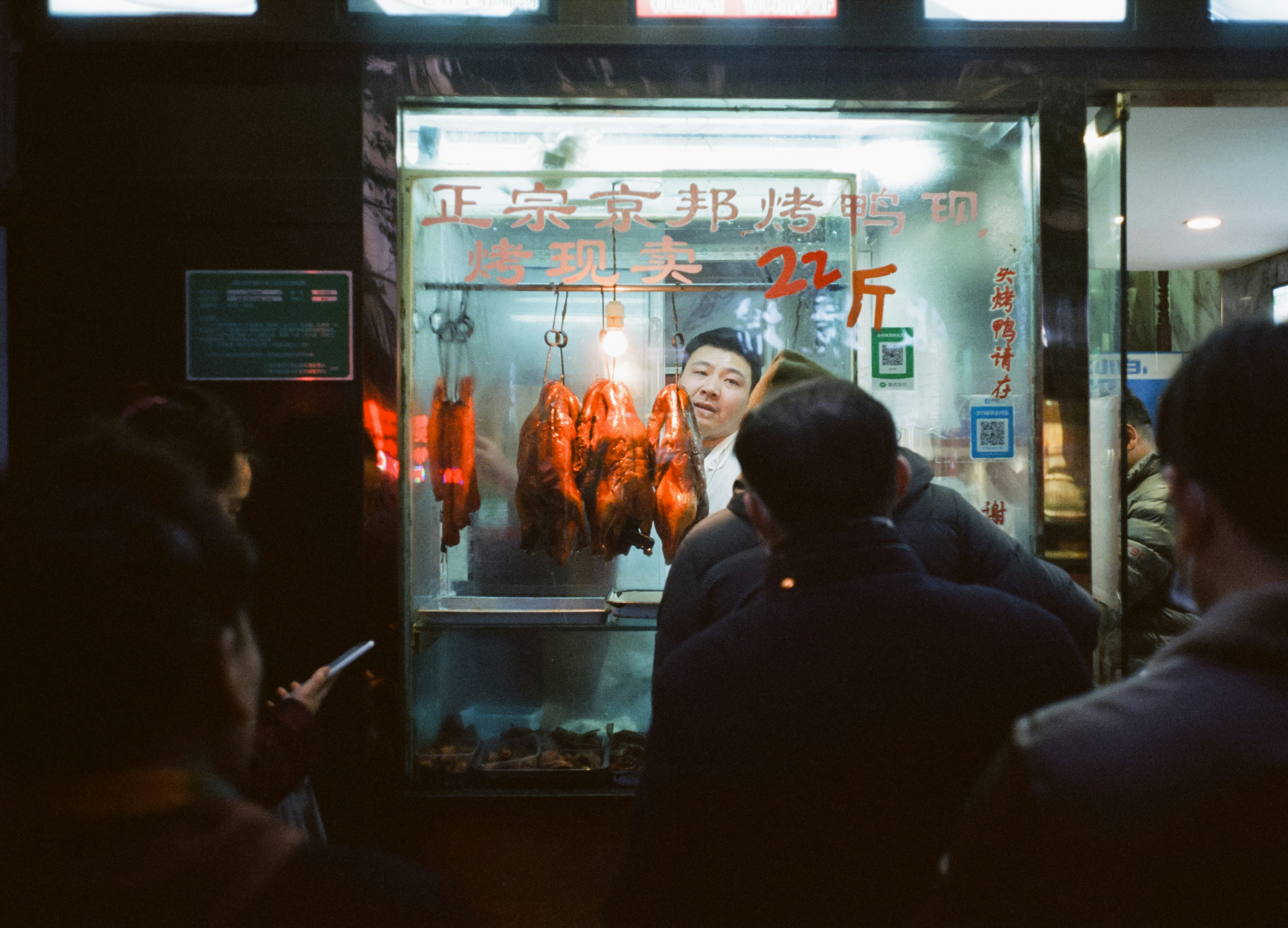 Vendor showcasing roasted meats in a bustling street market, with patrons eagerly observing. Neon lights illuminate the scene.