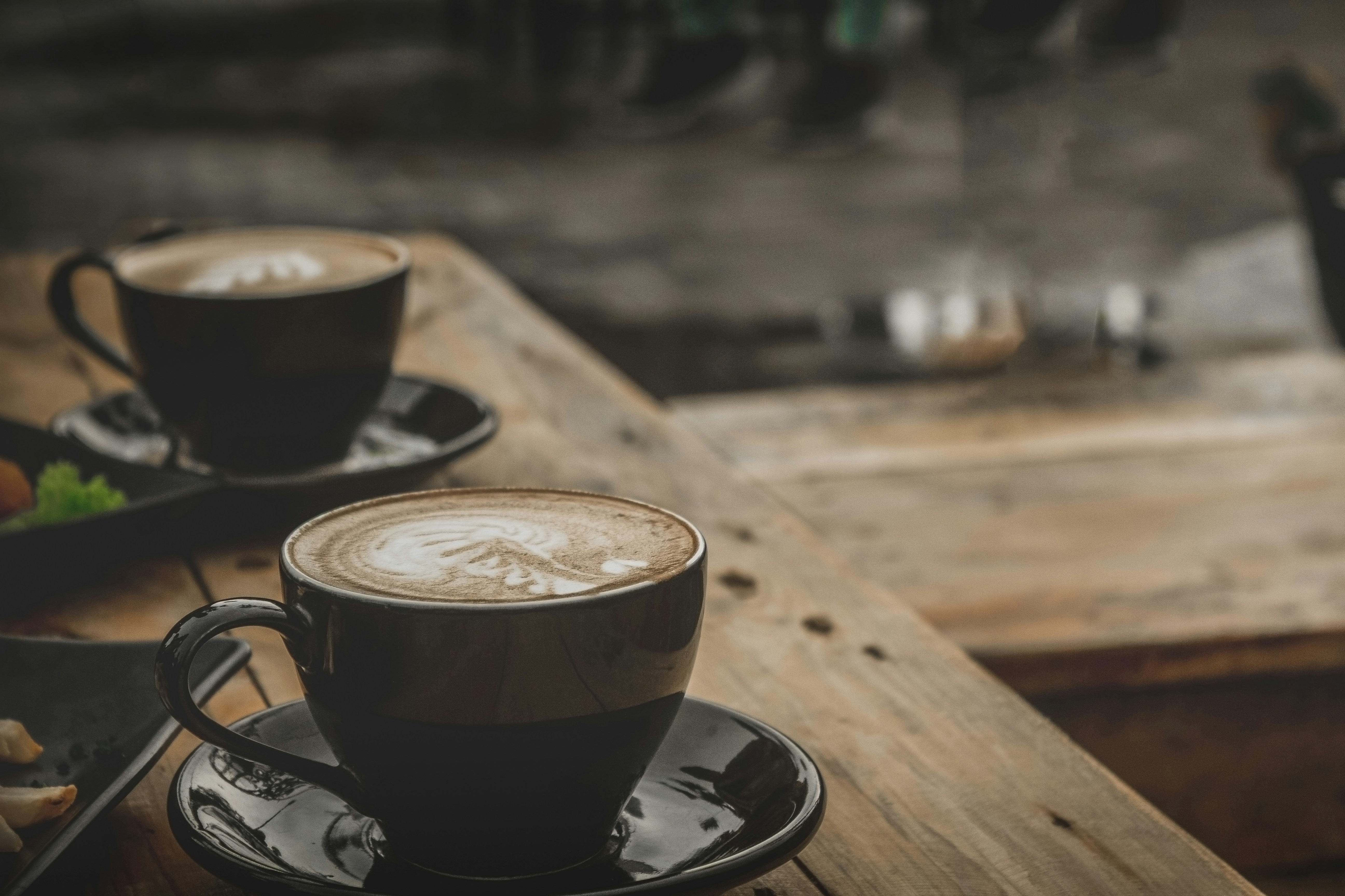 Two frothy cappuccinos in black cups on a wooden table, with a blurred rainy background.