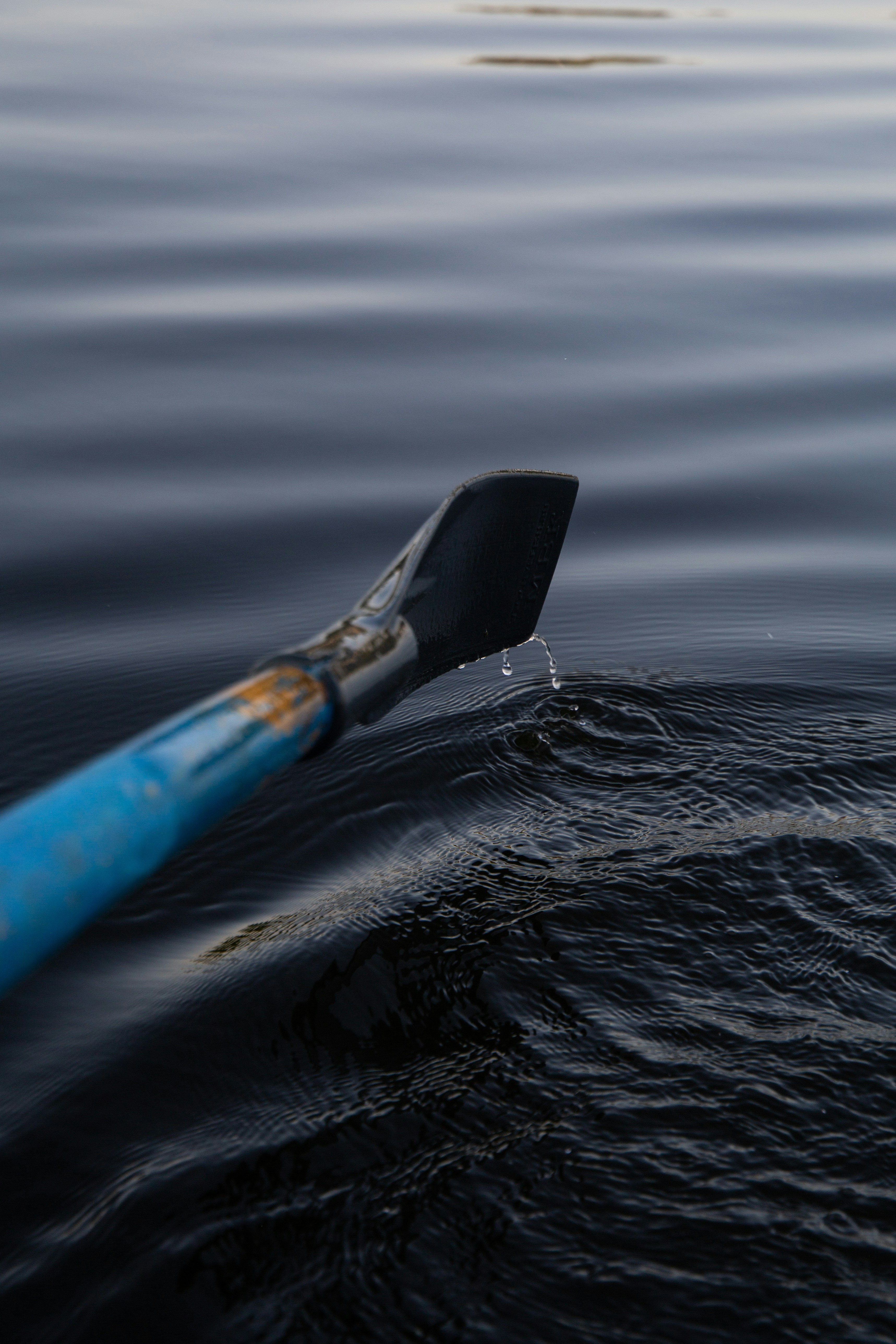 A close-up of a paddle dipping into tranquil water, creating gentle ripples. The scene captures the serene interaction between the paddle and the reflective surface.