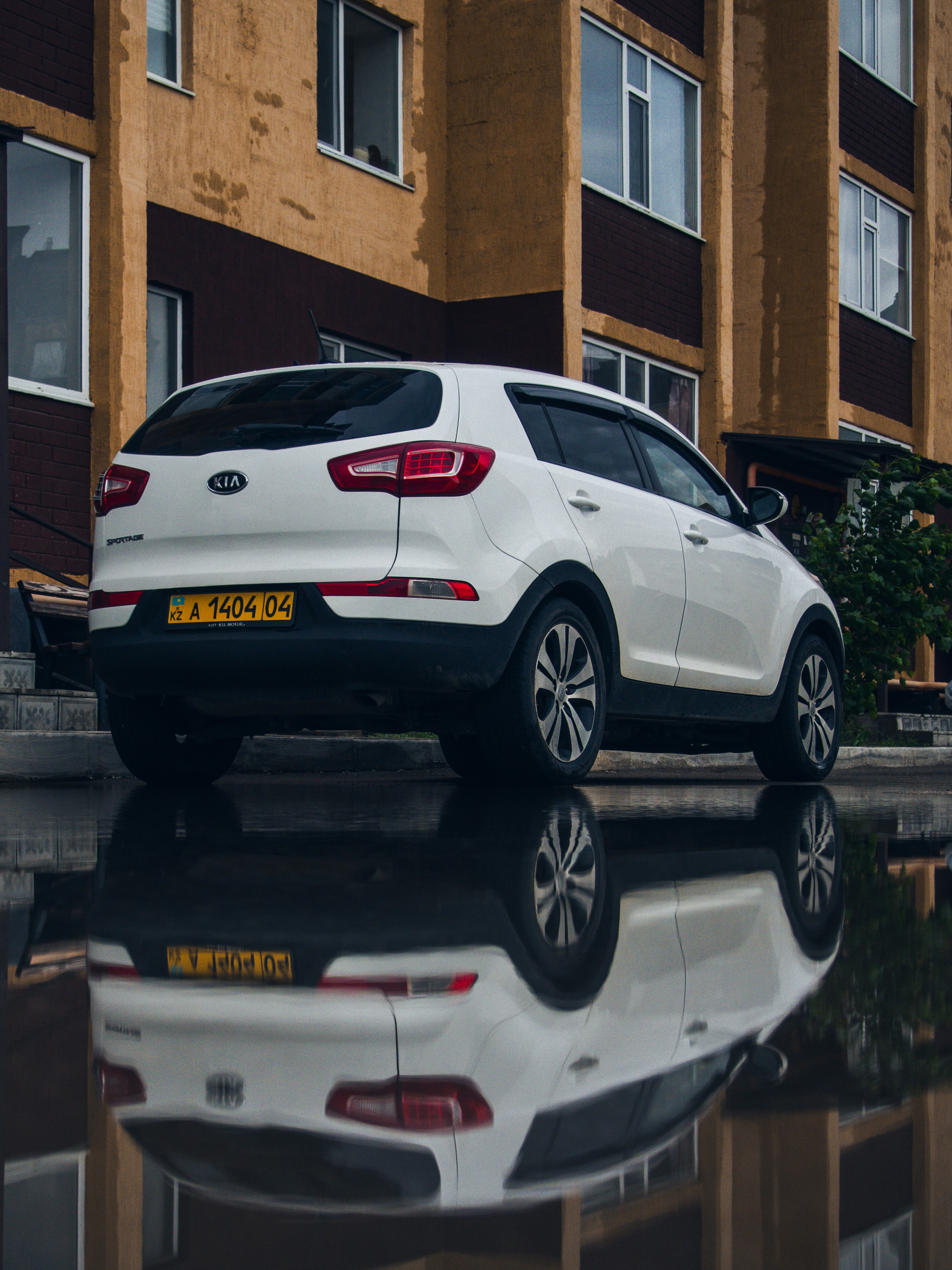 Rear view of a white Kia SUV parked beside a rain-washed street, its reflection clearly visible in a puddle against a mustard-yellow apartment block.