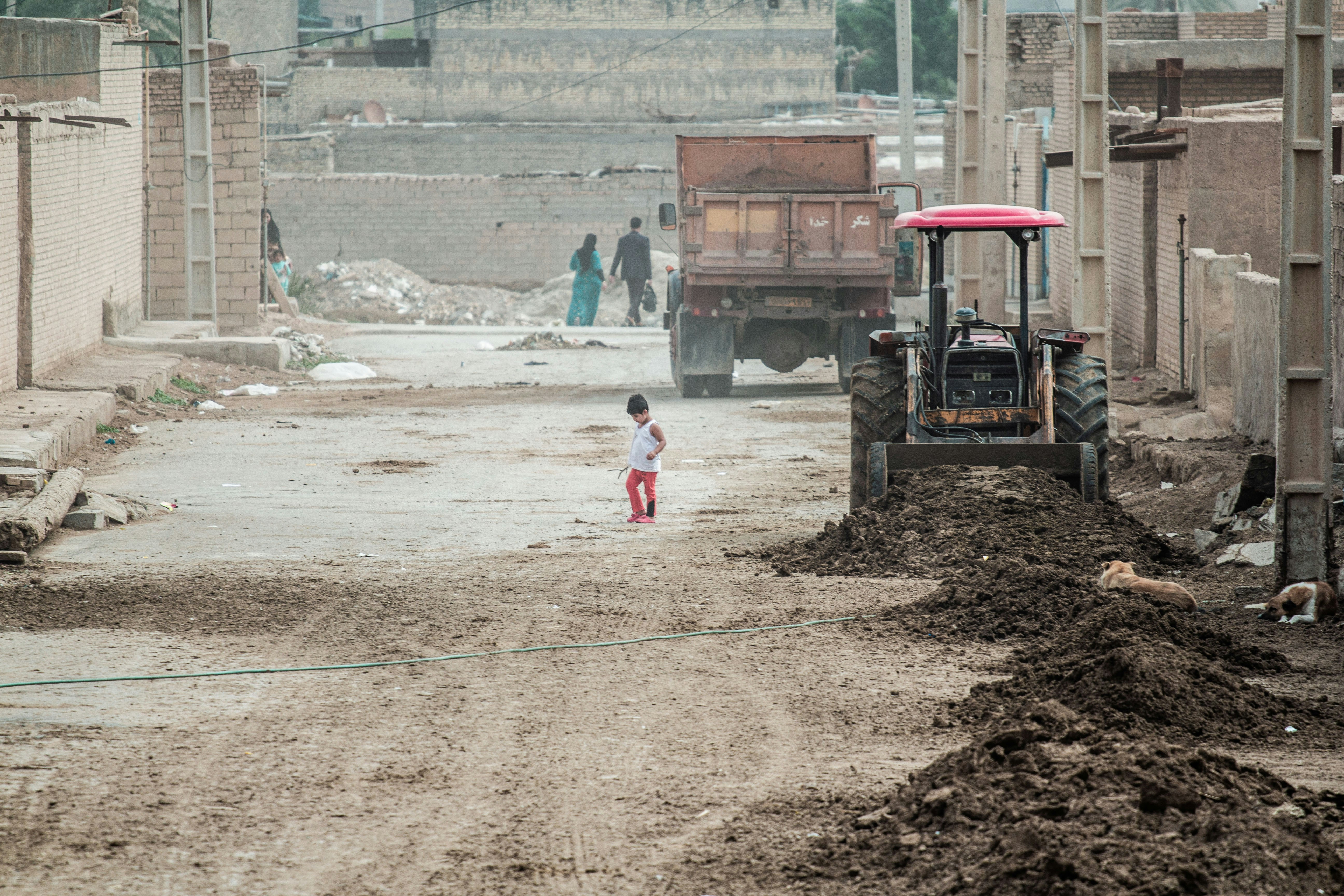 Child walking on a dusty street with construction vehicles and sleeping dogs nearby.