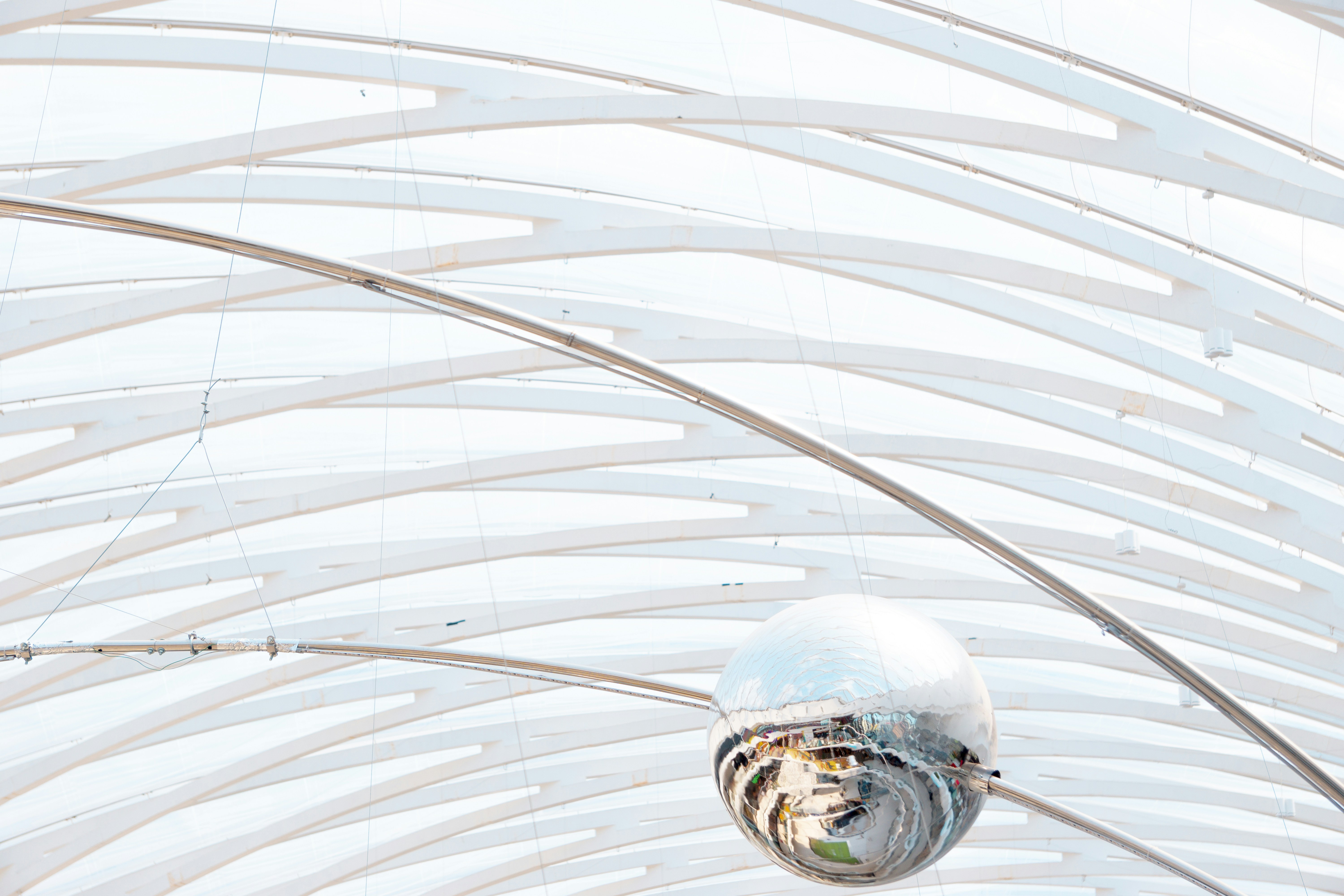 A polished metallic sphere hanging from a white, curved architectural structure, reflecting its surroundings. The interplay of light and shadow accentuates the design elements.