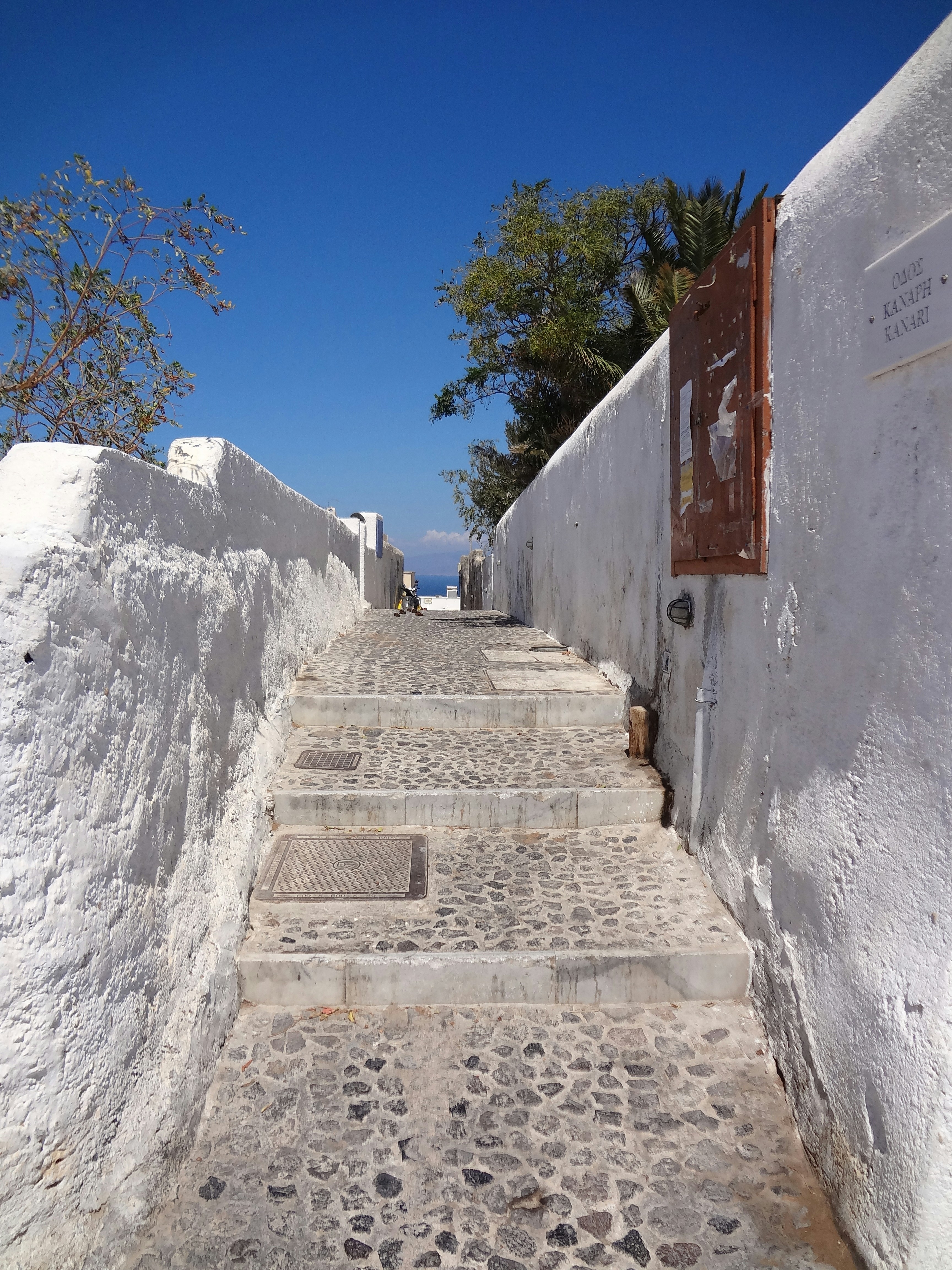 Narrow stone pathway bordered by whitewashed walls, leading towards a distant view of the sea under a clear blue sky.