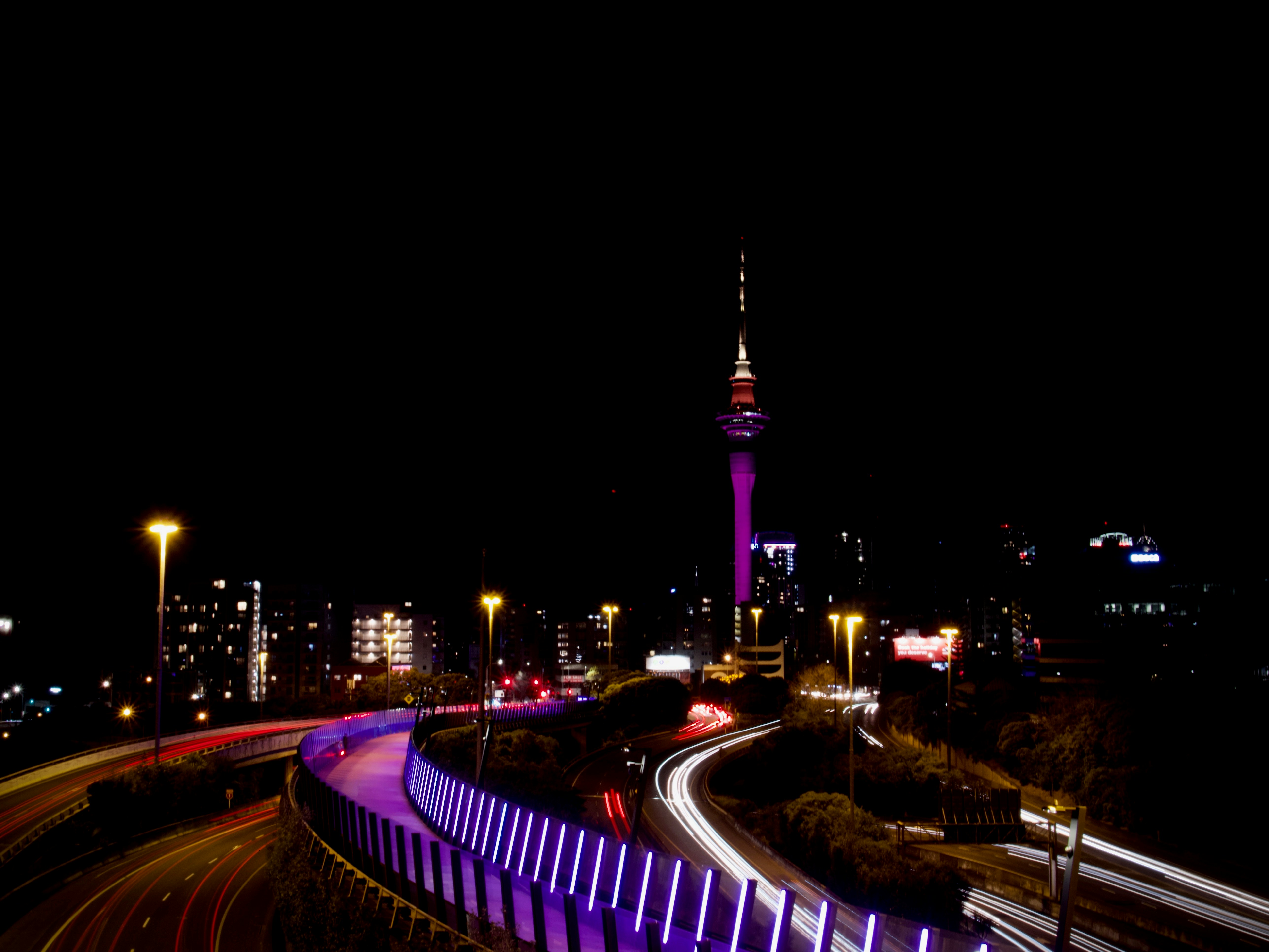 Vibrant cityscape illuminated by neon lights, featuring a prominent tower against the night sky. Dynamic light trails from moving vehicles enhance the scene.