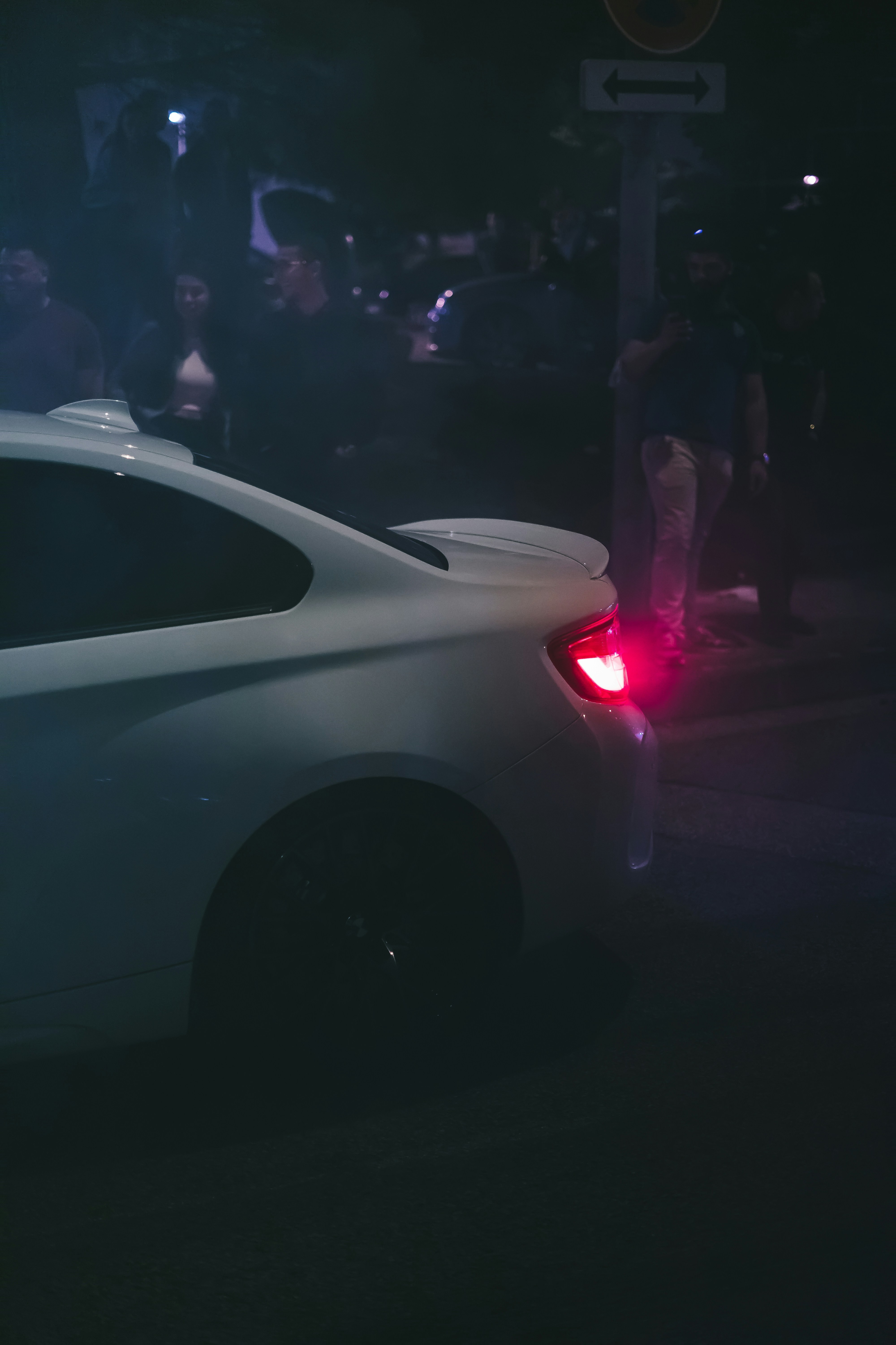 A sleek white car parked under dim street lights, with a glowing red tail light illuminating the surrounding shadows of an urban night.