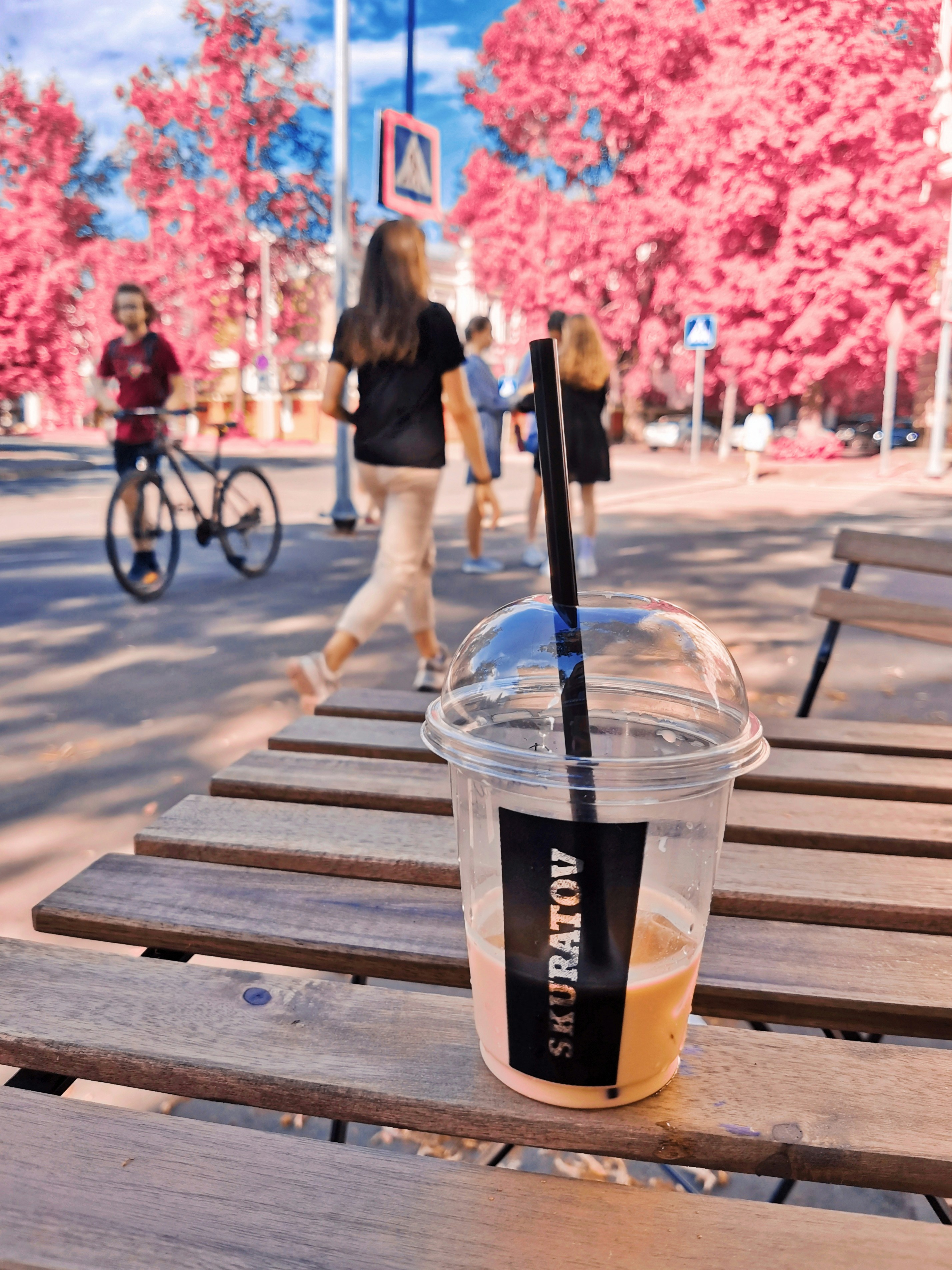 Coffee cup resting on a wooden table with vibrant pink trees and pedestrians in the background. The scene captures a lively urban atmosphere.