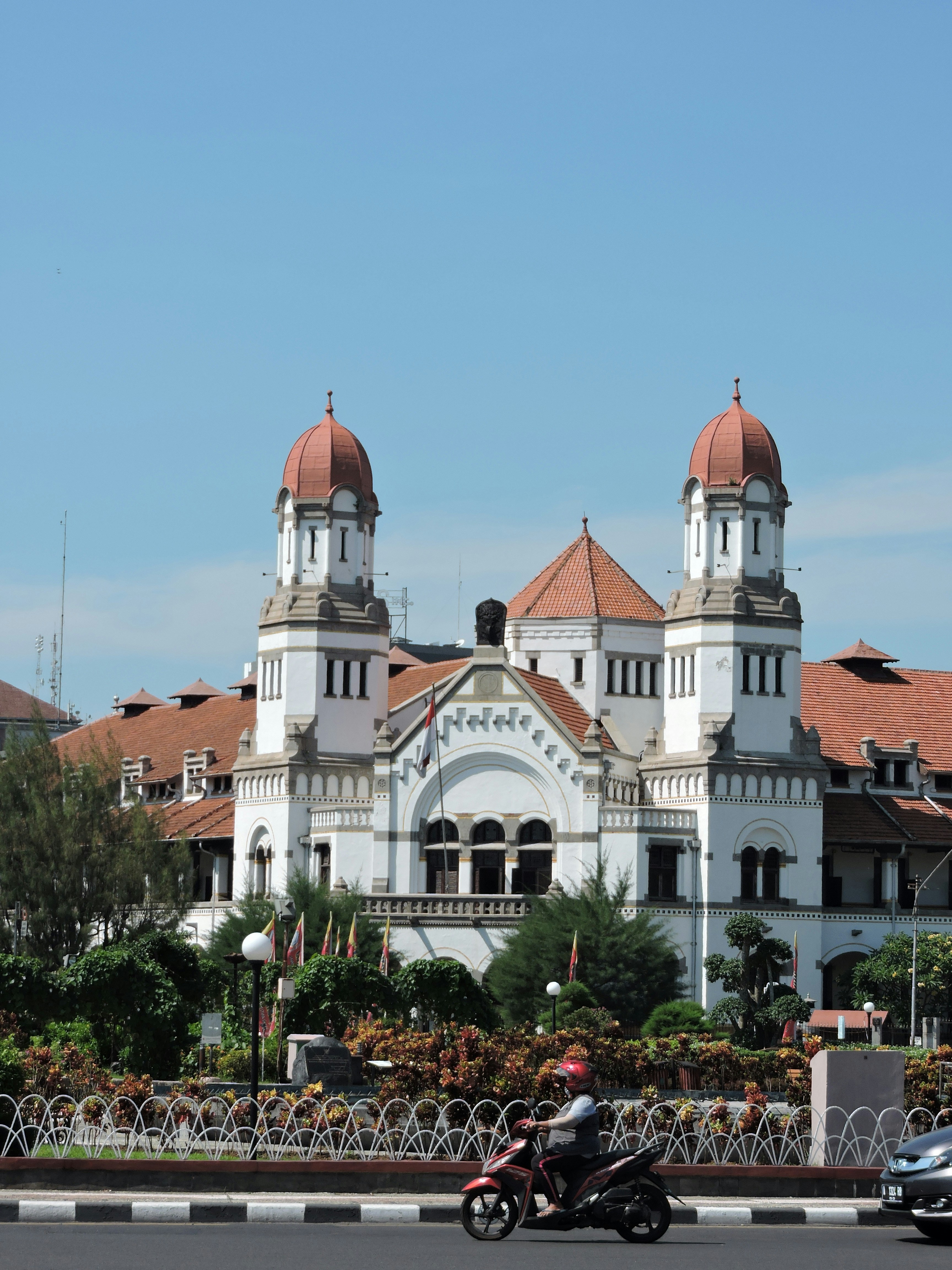 Historic building with prominent domes and intricate details surrounded by lush greenery and urban life.