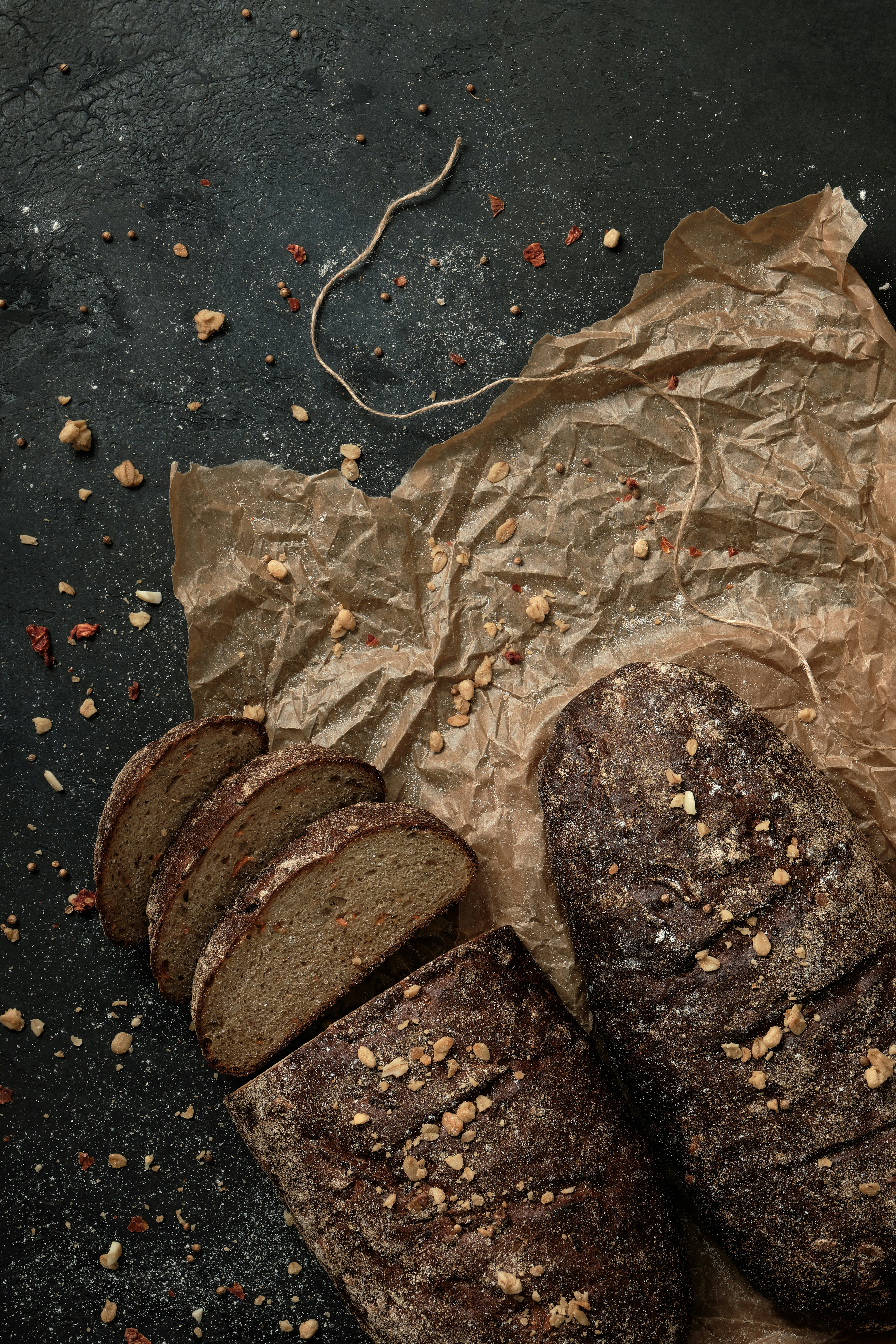 Two rustic loaves of bread on crinkled parchment, with slices fanned out and scattered nuts and spices around them.