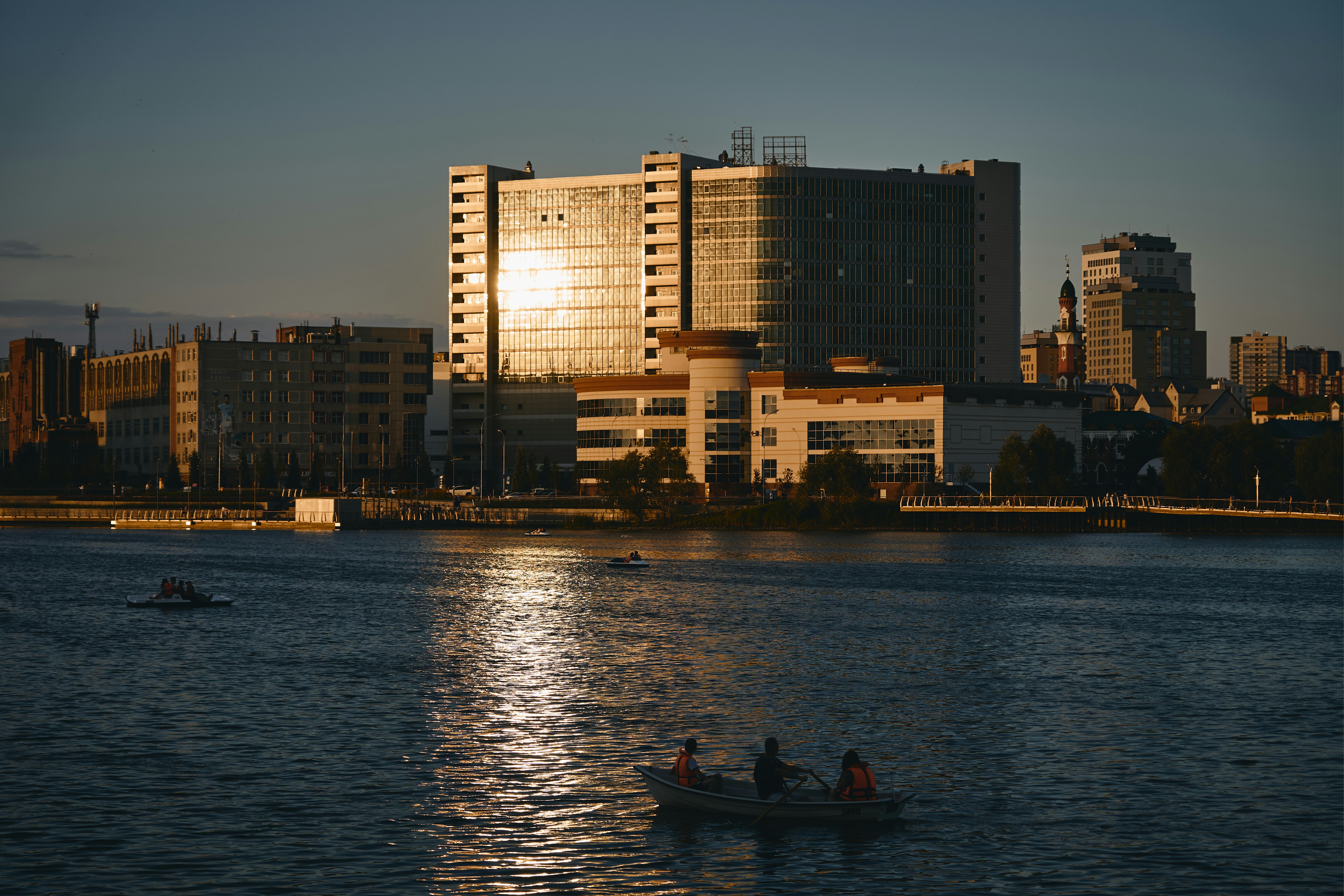 Three individuals in a small boat navigate a shimmering river, with a modern building reflecting the evening light in the background.