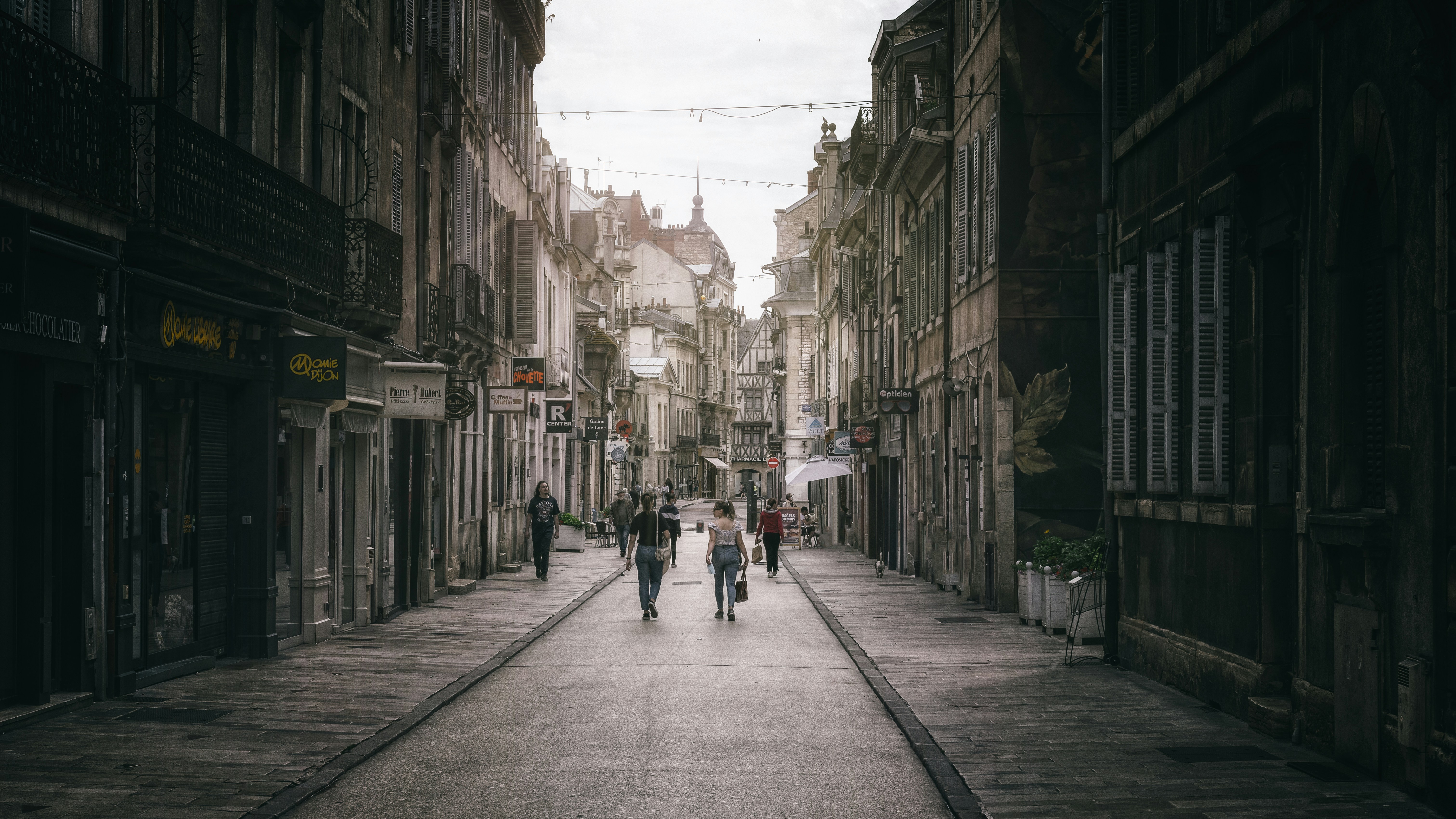 Two people walking down a narrow cobblestone street flanked by old buildings under a cloudy sky.