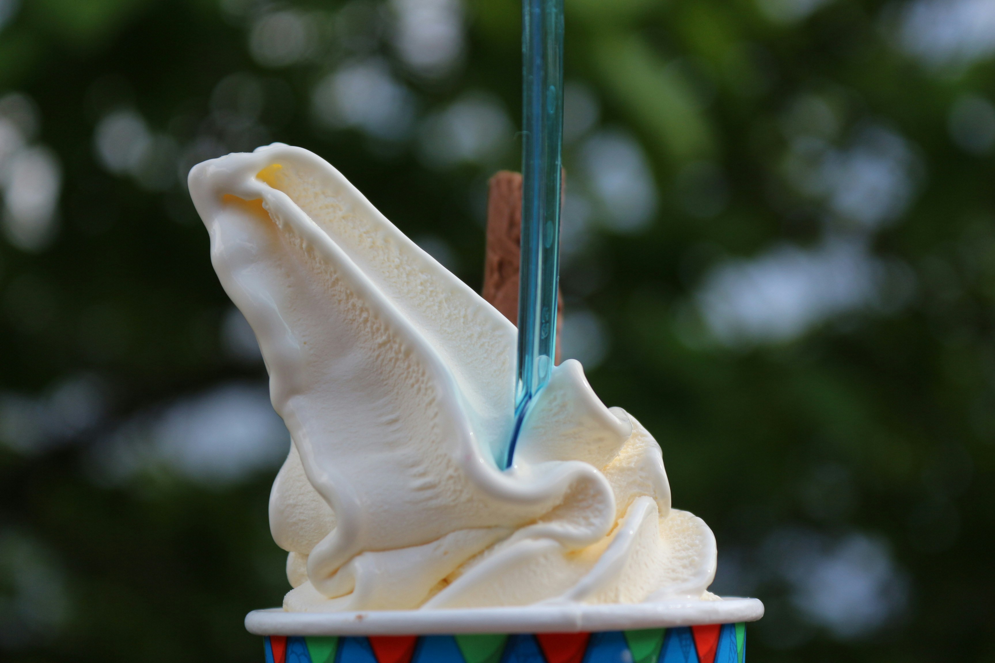 A swirl of creamy soft serve ice cream topped with a blue straw and a wooden stick, set against a blurred green backdrop.