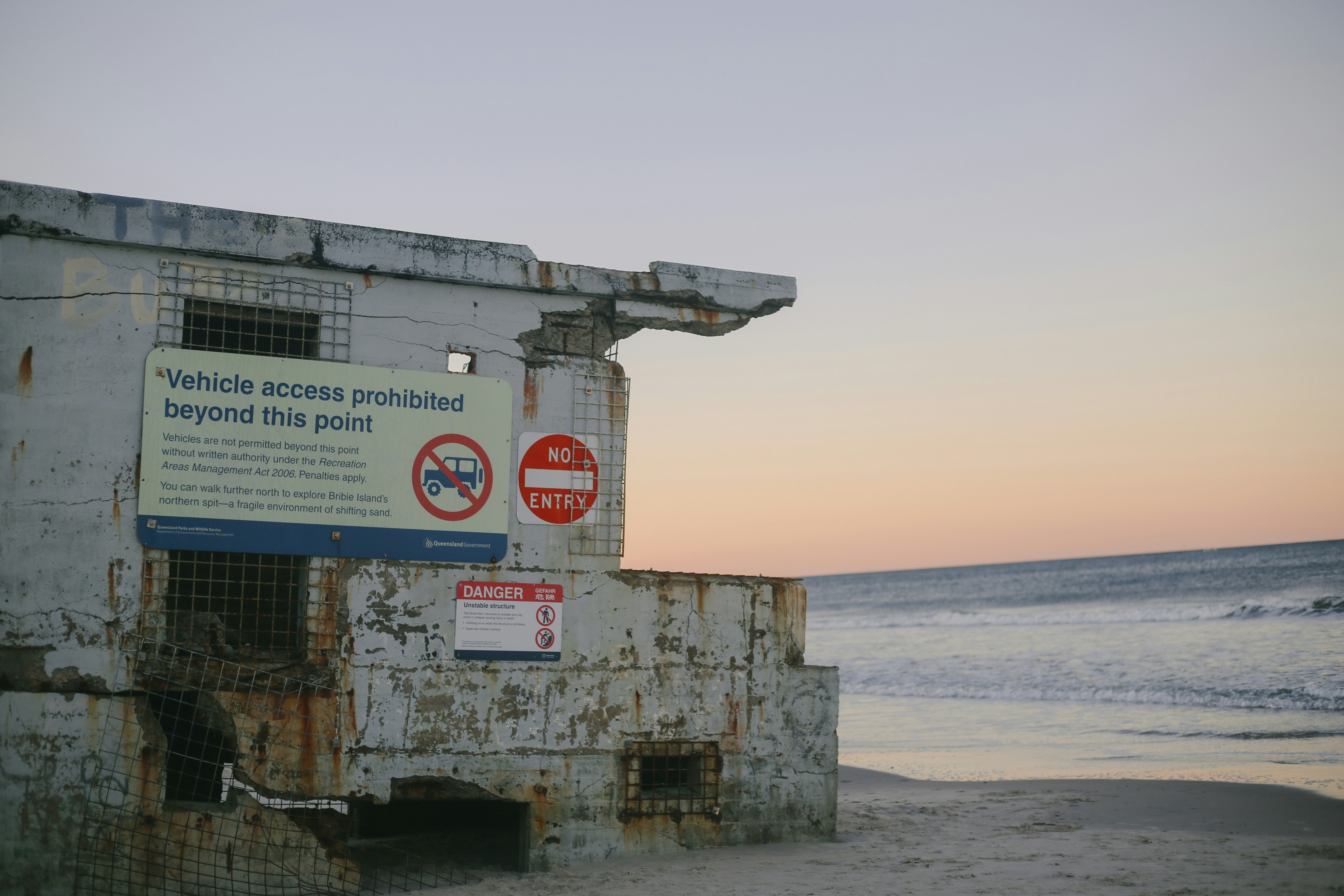 Weathered signpost warning against vehicle access, standing on a beach at dusk. The fading light highlights the textures of the structure and surrounding waves.