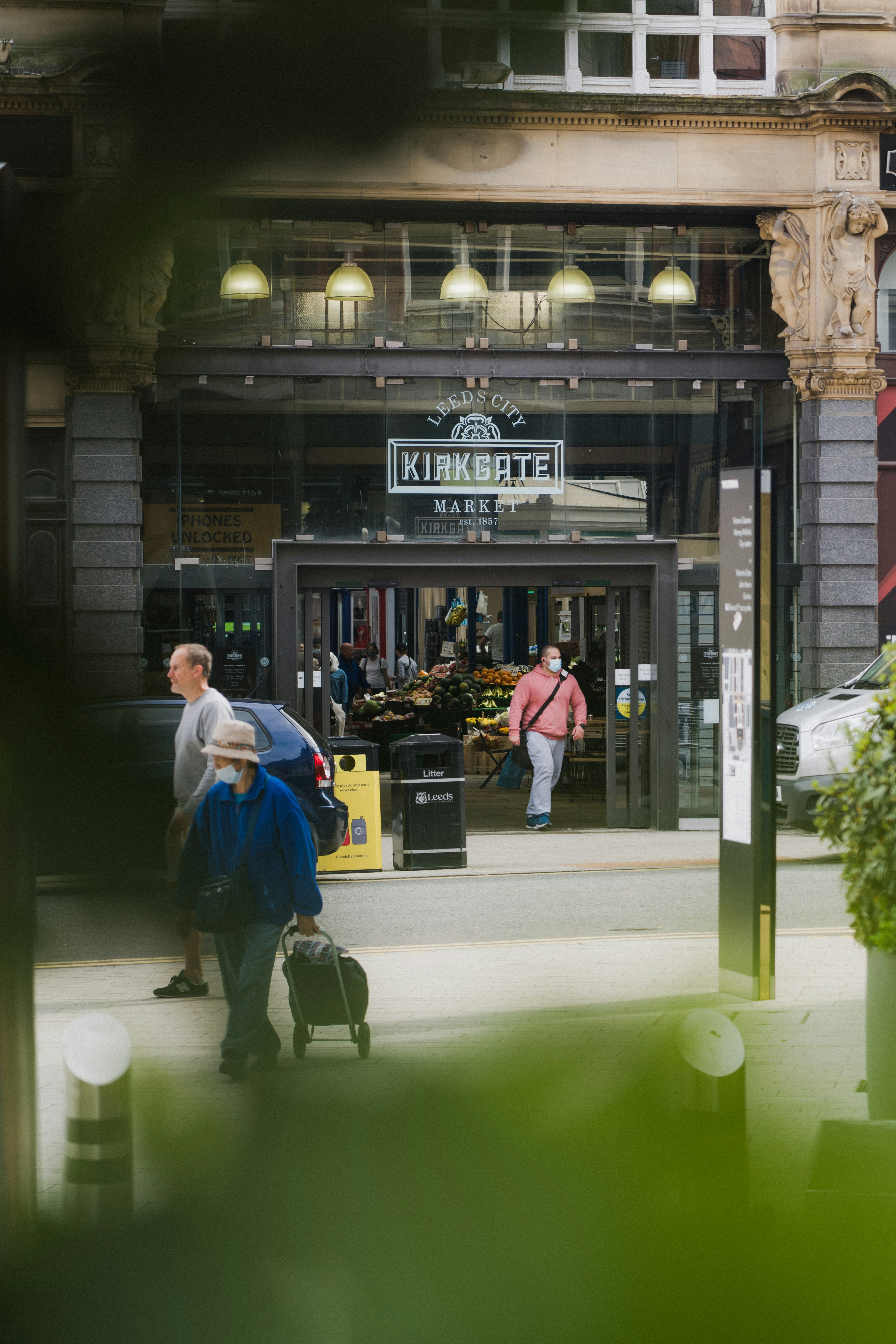 A vibrant scene at Kirkgate Market showcasing shoppers and fresh produce, framed by lush greenery in the foreground.