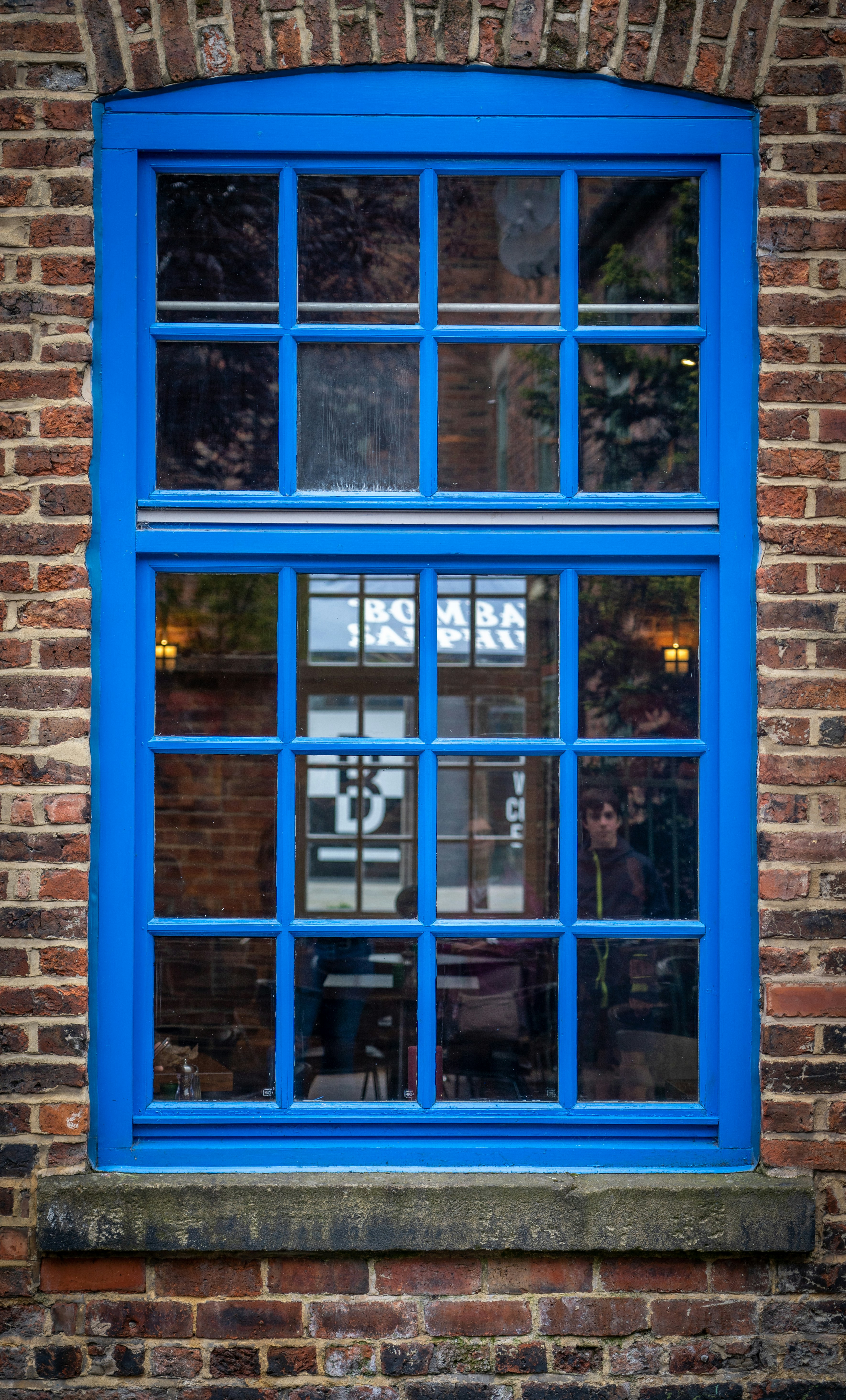A vibrant blue window frame reflects the lively atmosphere of a café, showcasing the interplay of light and shadow. The scene captures a moment of urban life.