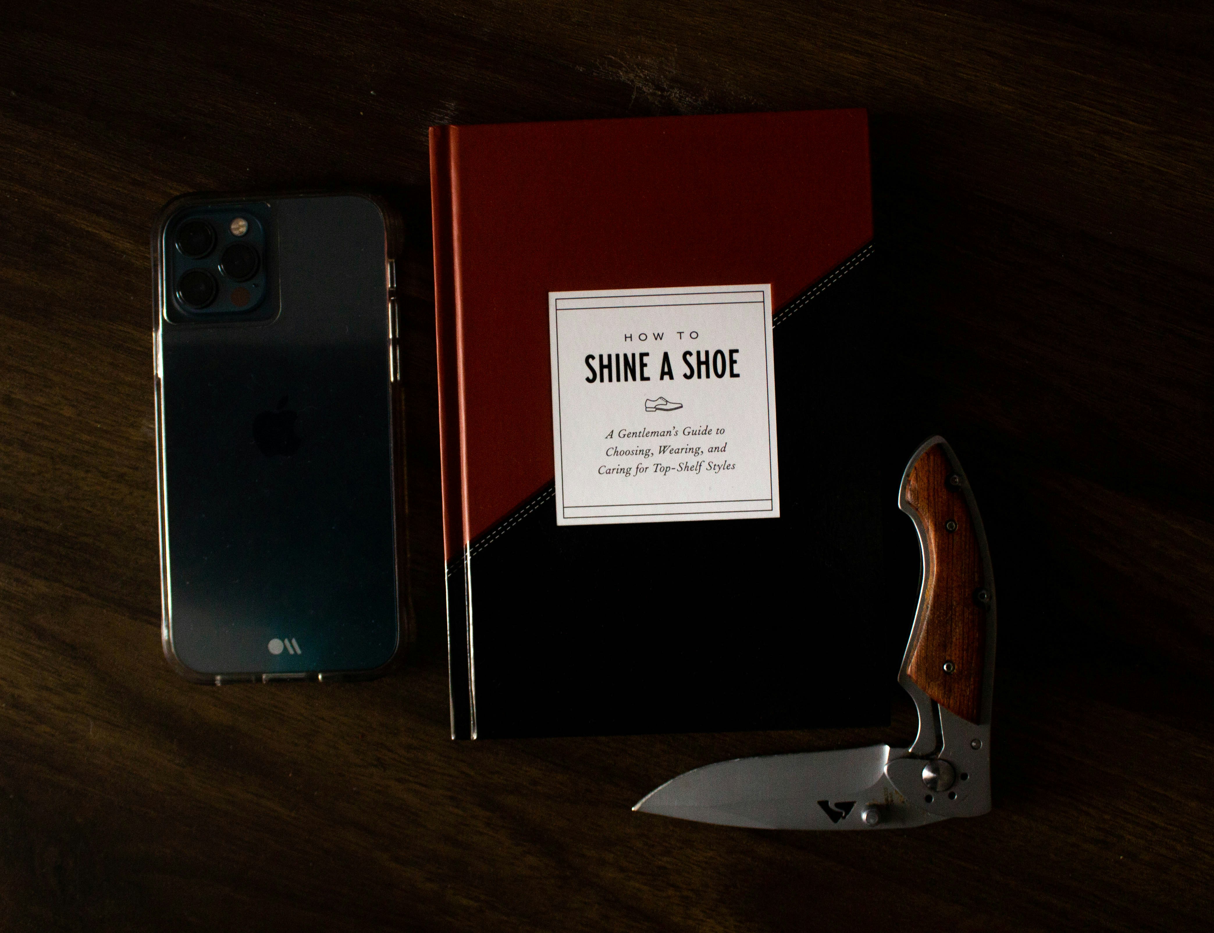 A stylish book titled 'How to Shine a Shoe' lies next to a smartphone and a folding knife on a wooden surface.