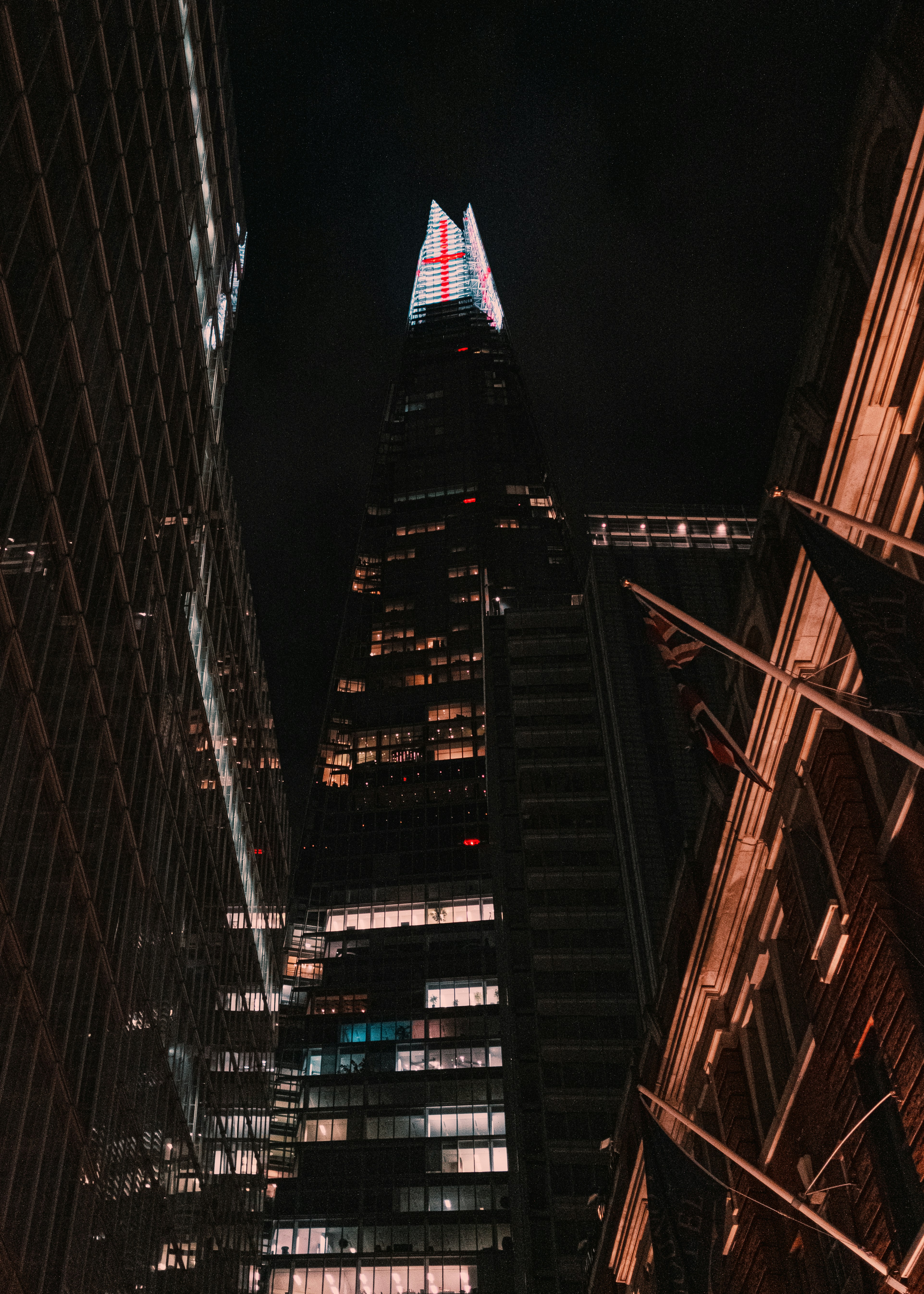 Illuminated skyscraper towering amidst reflective glass buildings under a night sky. The structure's apex is highlighted with striking lights.