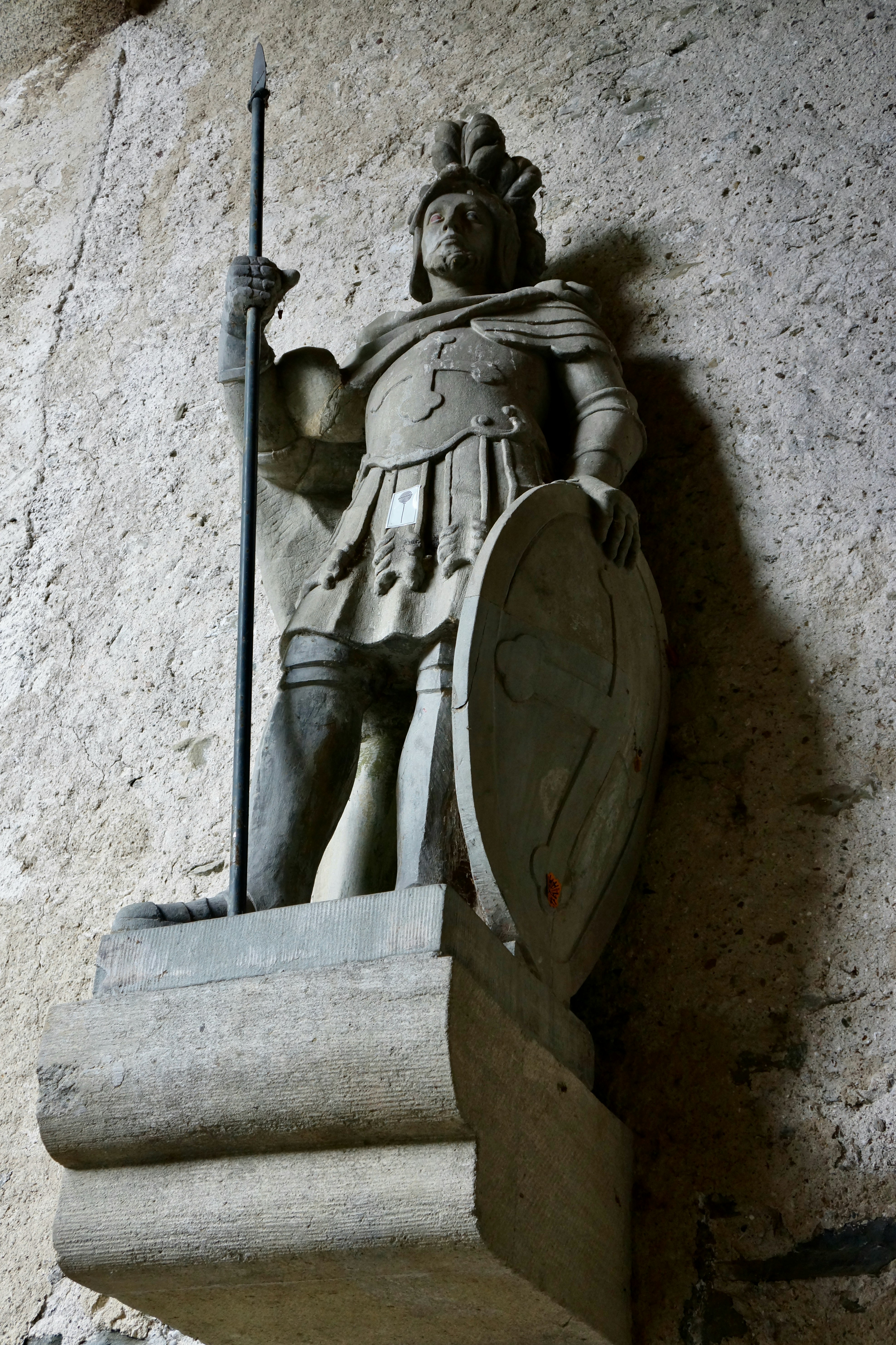 Stone statue of a Roman warrior holding a spear and shield, mounted against a textured wall.