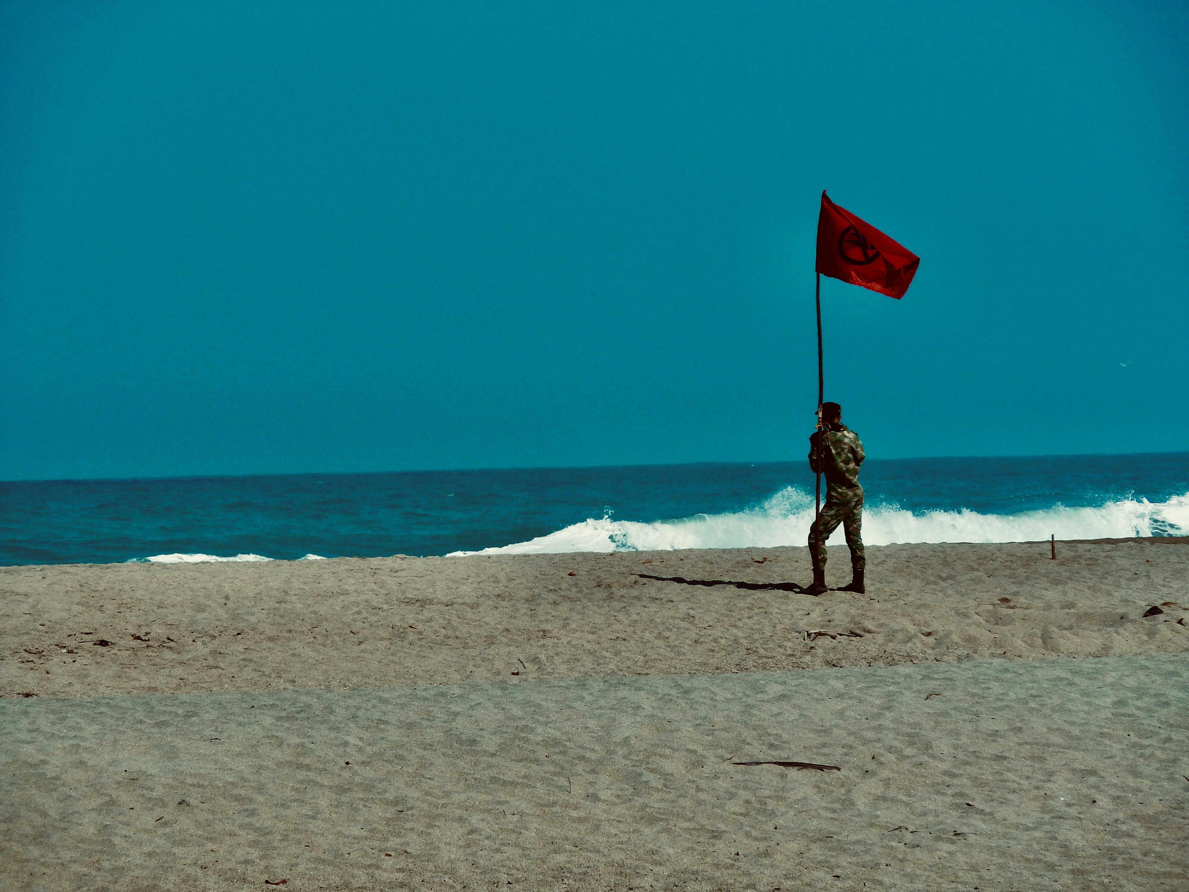 Soldier standing on sandy beach, proudly holding a red flag against a backdrop of ocean waves and clear blue sky.