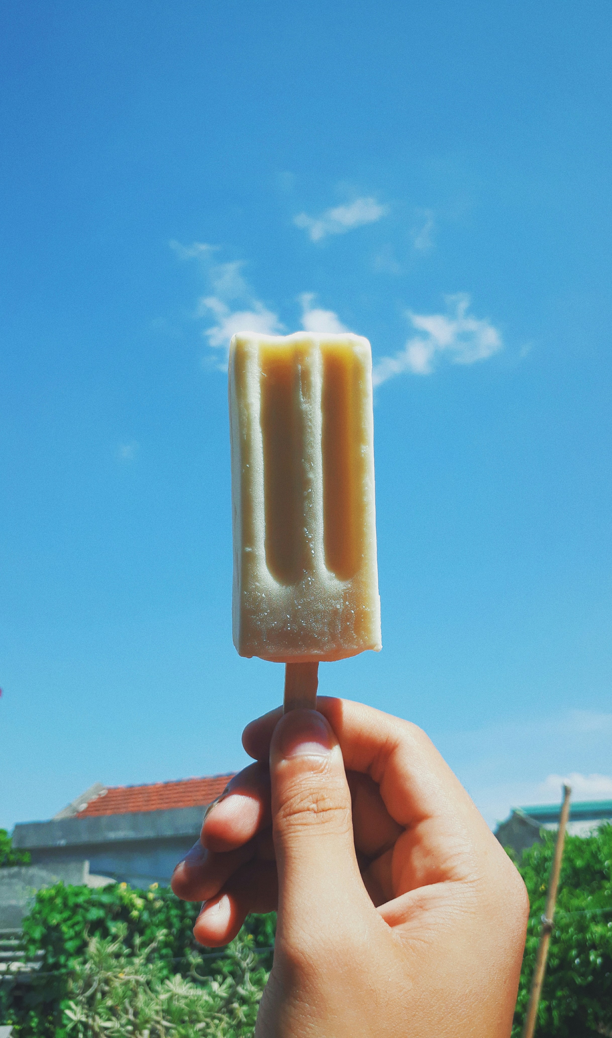 Hand holding a creamy popsicle against a bright blue sky with wispy clouds.