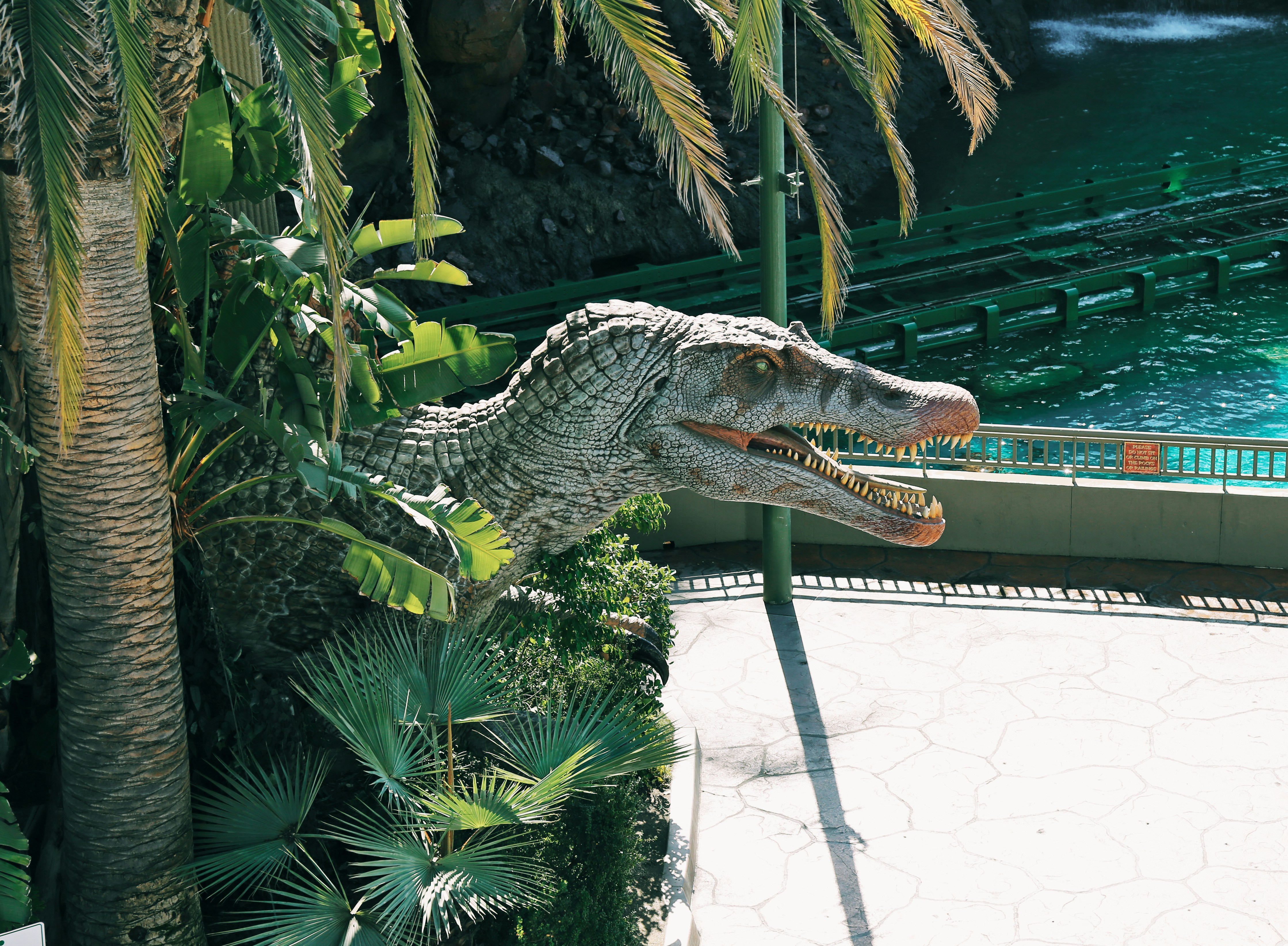 A detailed dinosaur sculpture emerges from lush greenery, set against a vibrant blue water backdrop. The scene evokes a prehistoric ambiance.