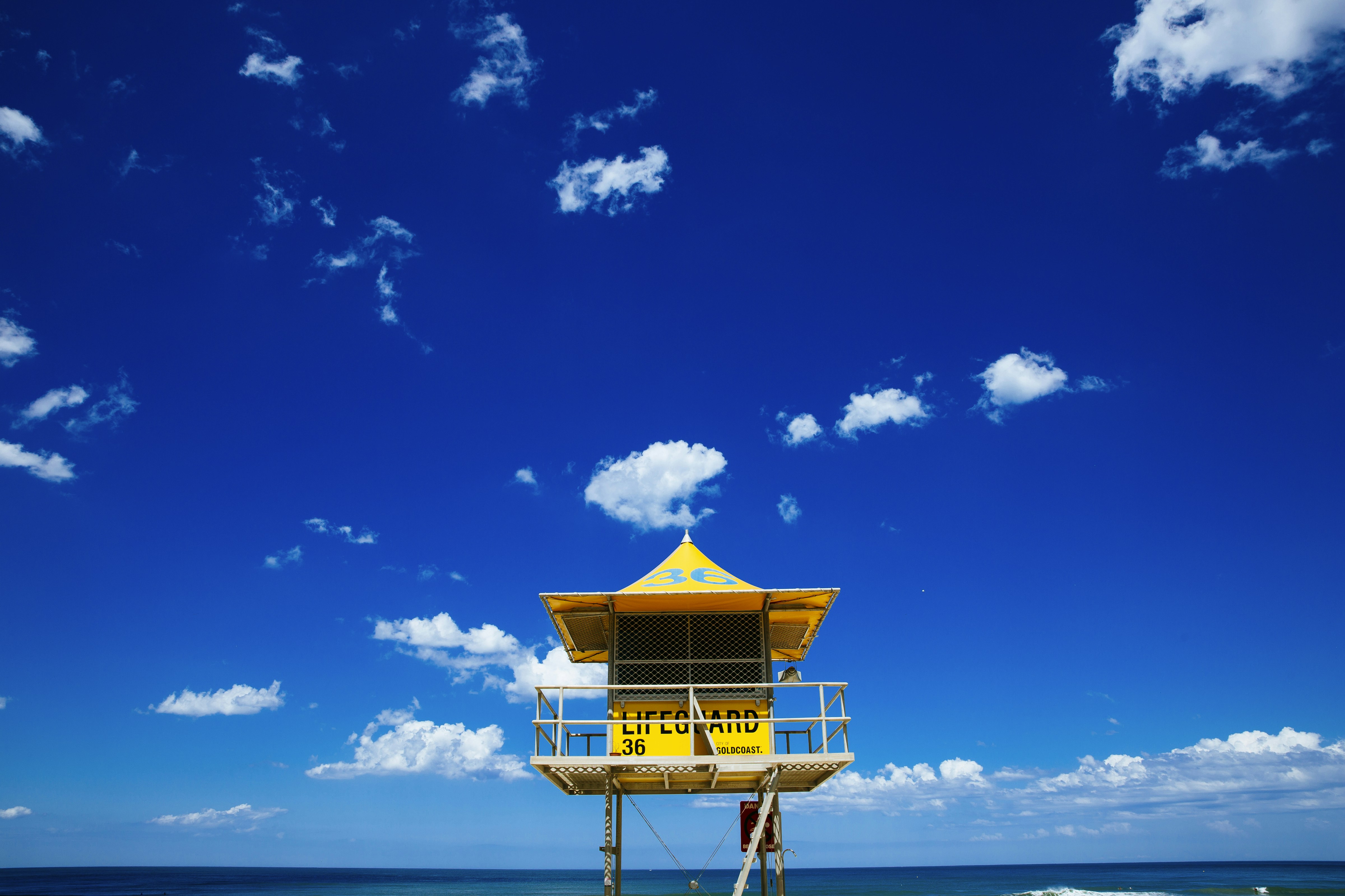 Bright yellow lifeguard tower stands against a vivid blue sky dotted with fluffy clouds.