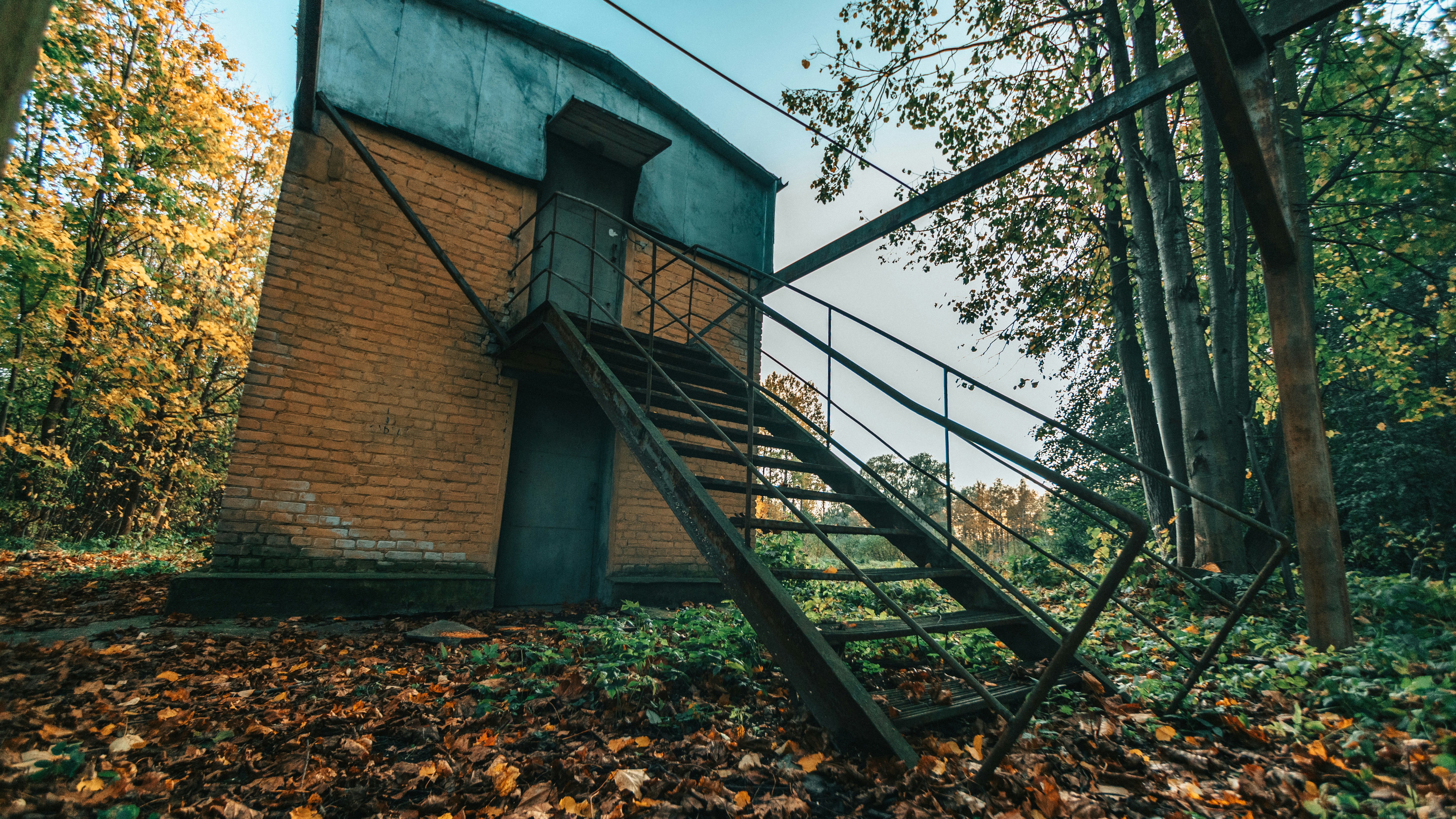 An old brick utility building with a metal staircase at the Pulkovo Observatory, surrounded by autumn trees and fallen leaves. The muted colors and weathered textures evoke a quiet, nostalgic atmosphere of scientific history.