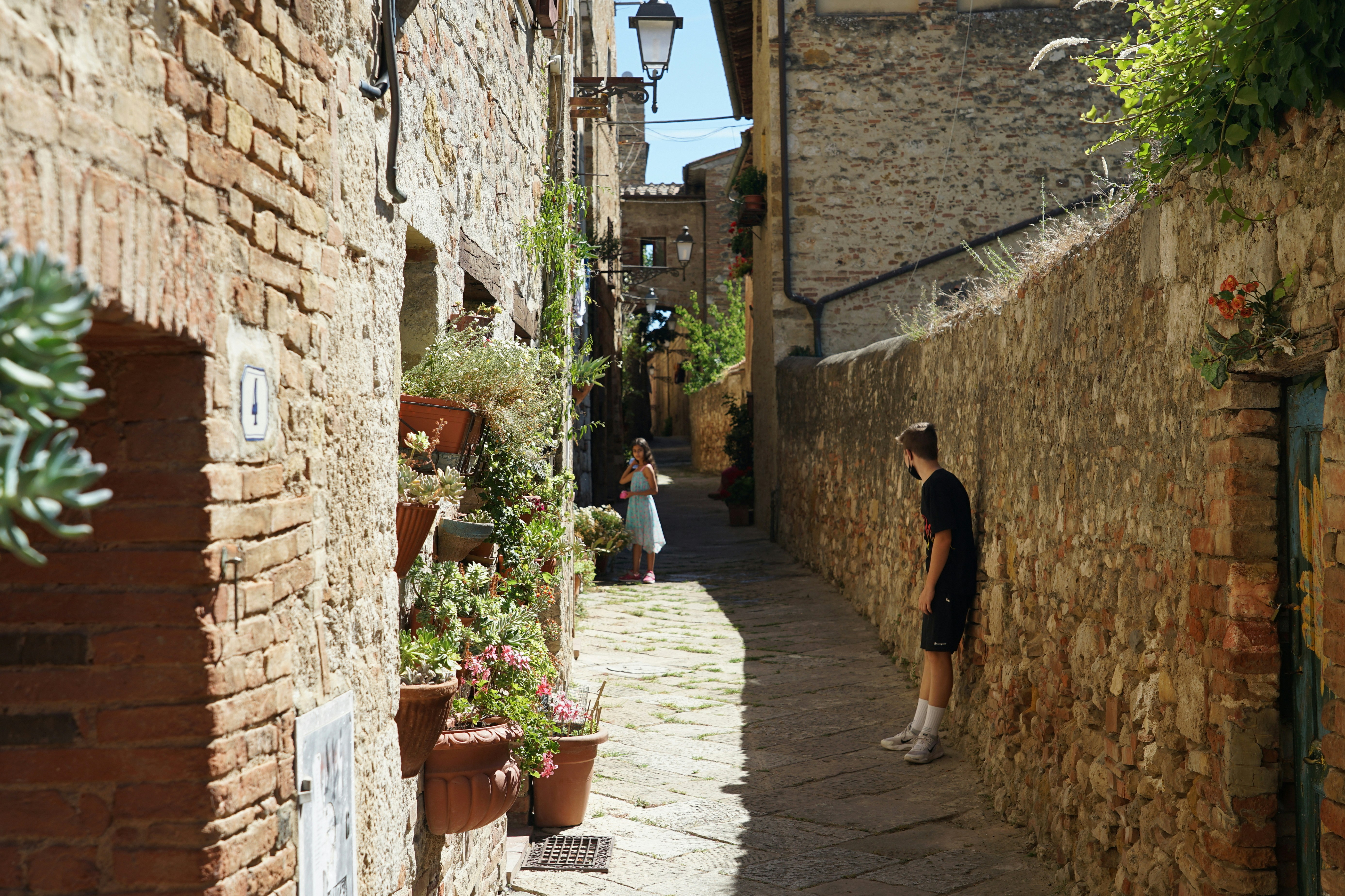 Narrow stone alleyway in Colle di Val d'Elsa with potted plants and rustic architecture under bright daylight.