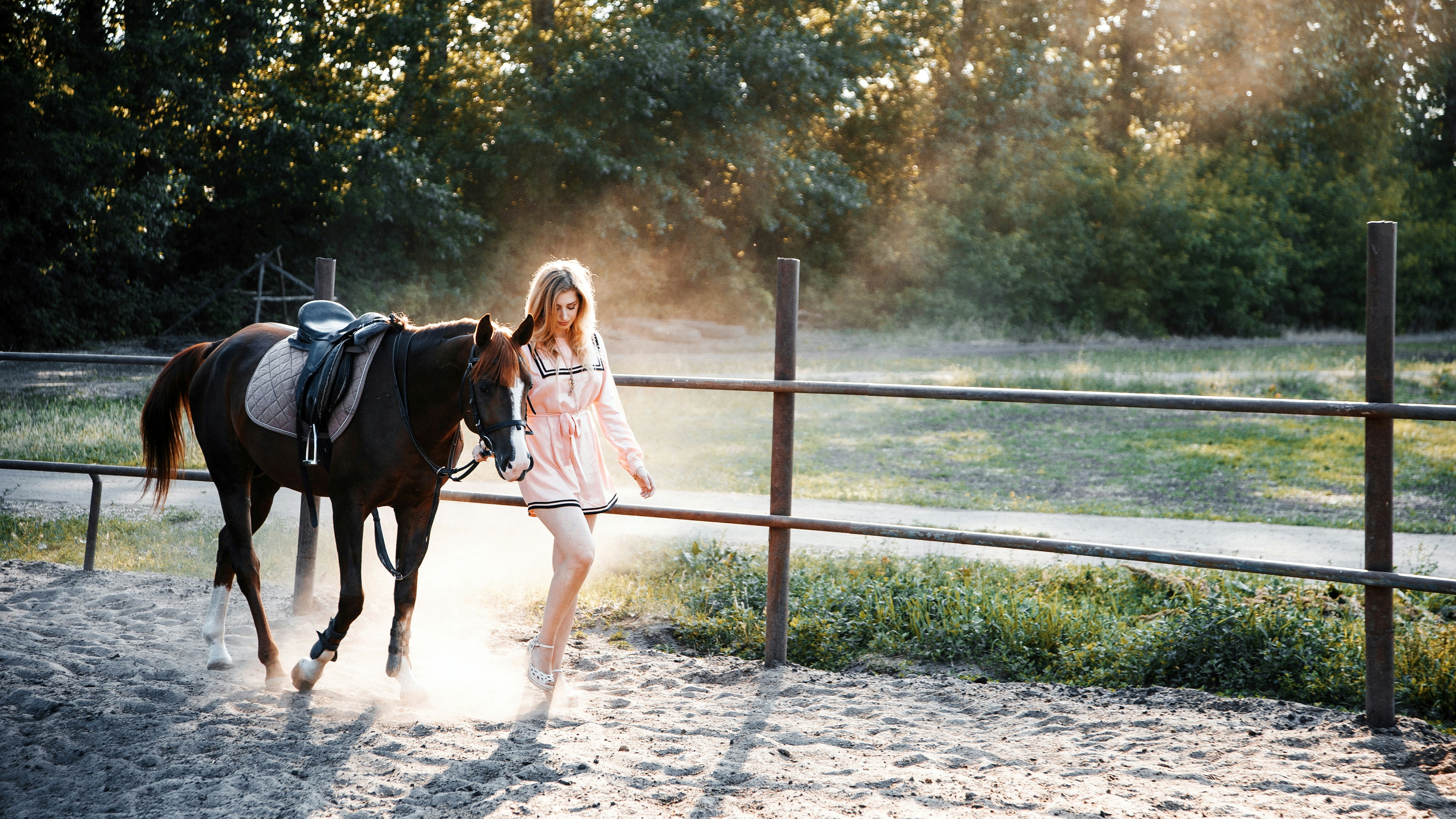 A woman in a flowing dress walks alongside a horse in a sunlit arena, dust swirling around them. The serene atmosphere captures a moment of connection between rider and steed.