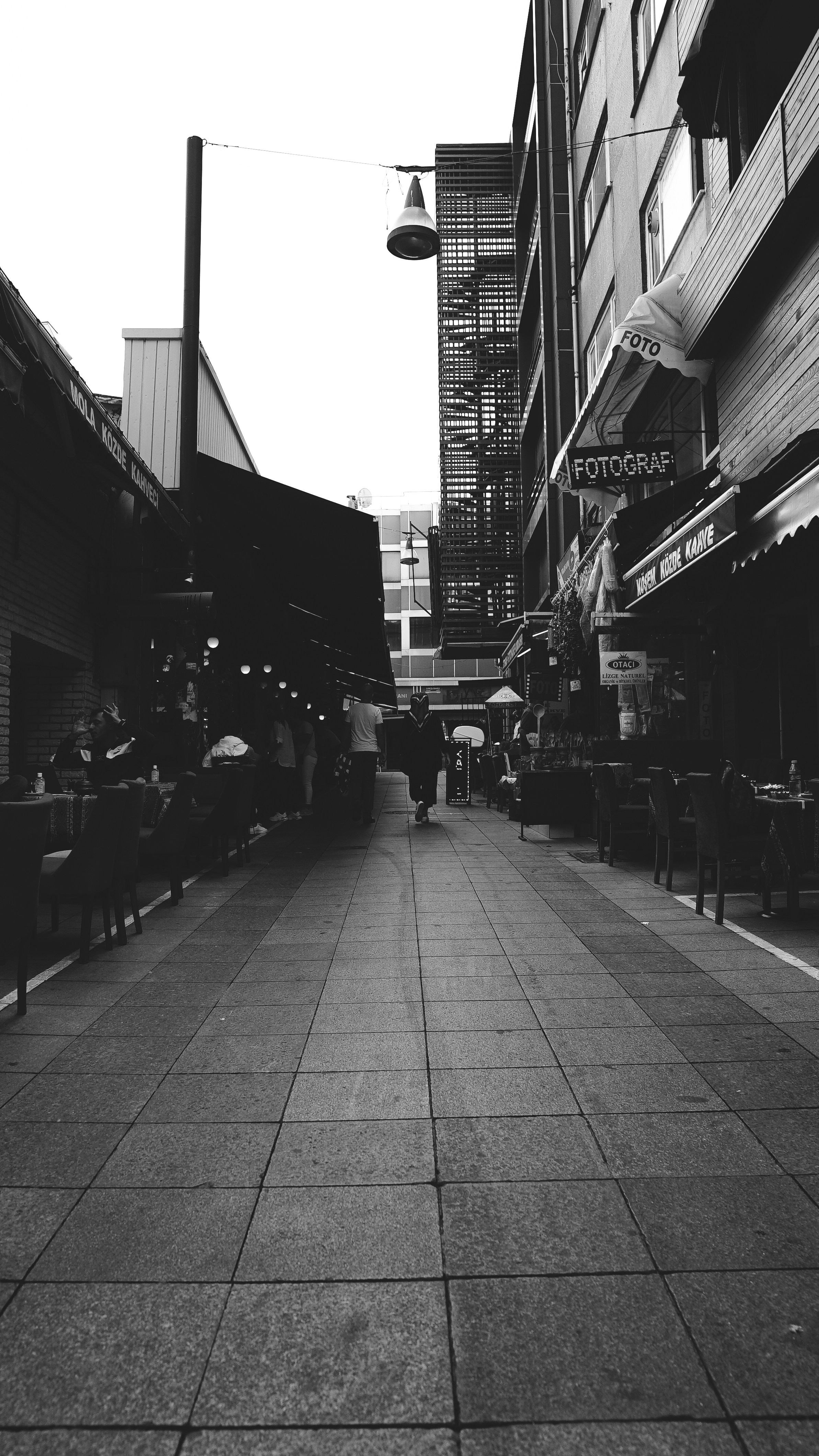 Narrow alleyway lined with outdoor seating and storefronts, leading towards a distant figure under a muted sky.