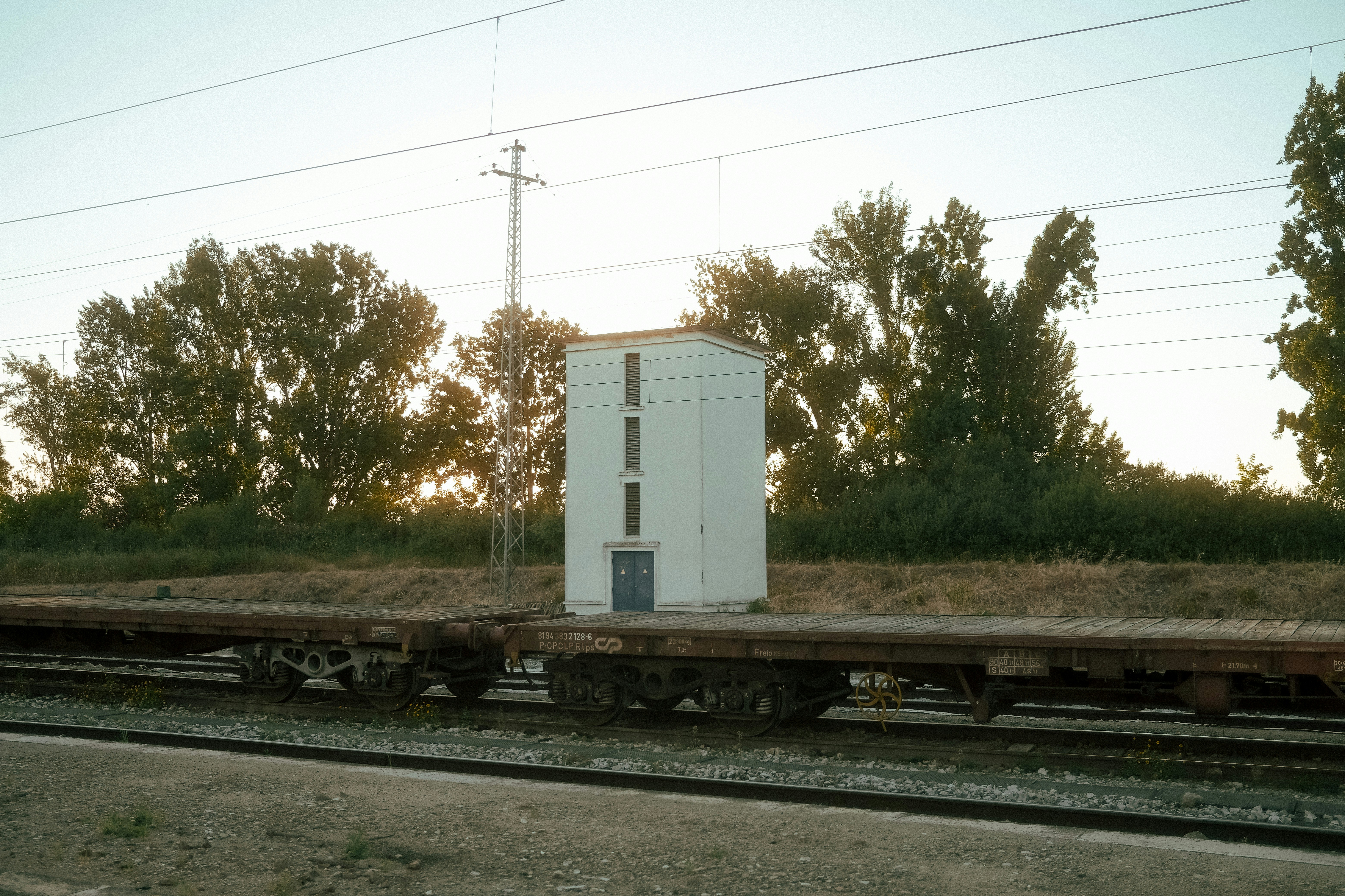 A stark white structure positioned on a train flatcar, surrounded by greenery and power lines at dusk.