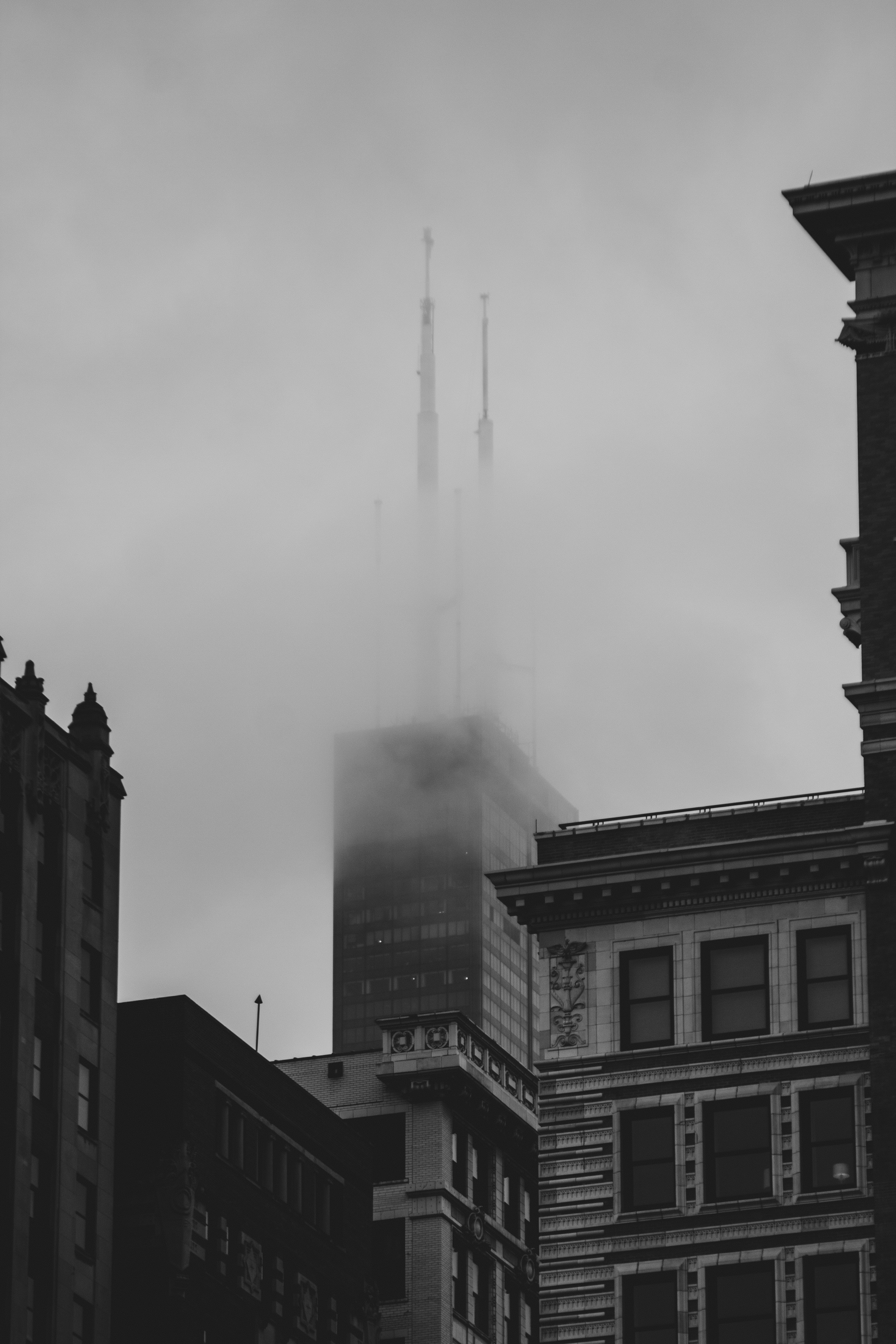 Tall skyscraper shrouded in fog, partially obscured by surrounding buildings in a monochrome setting.